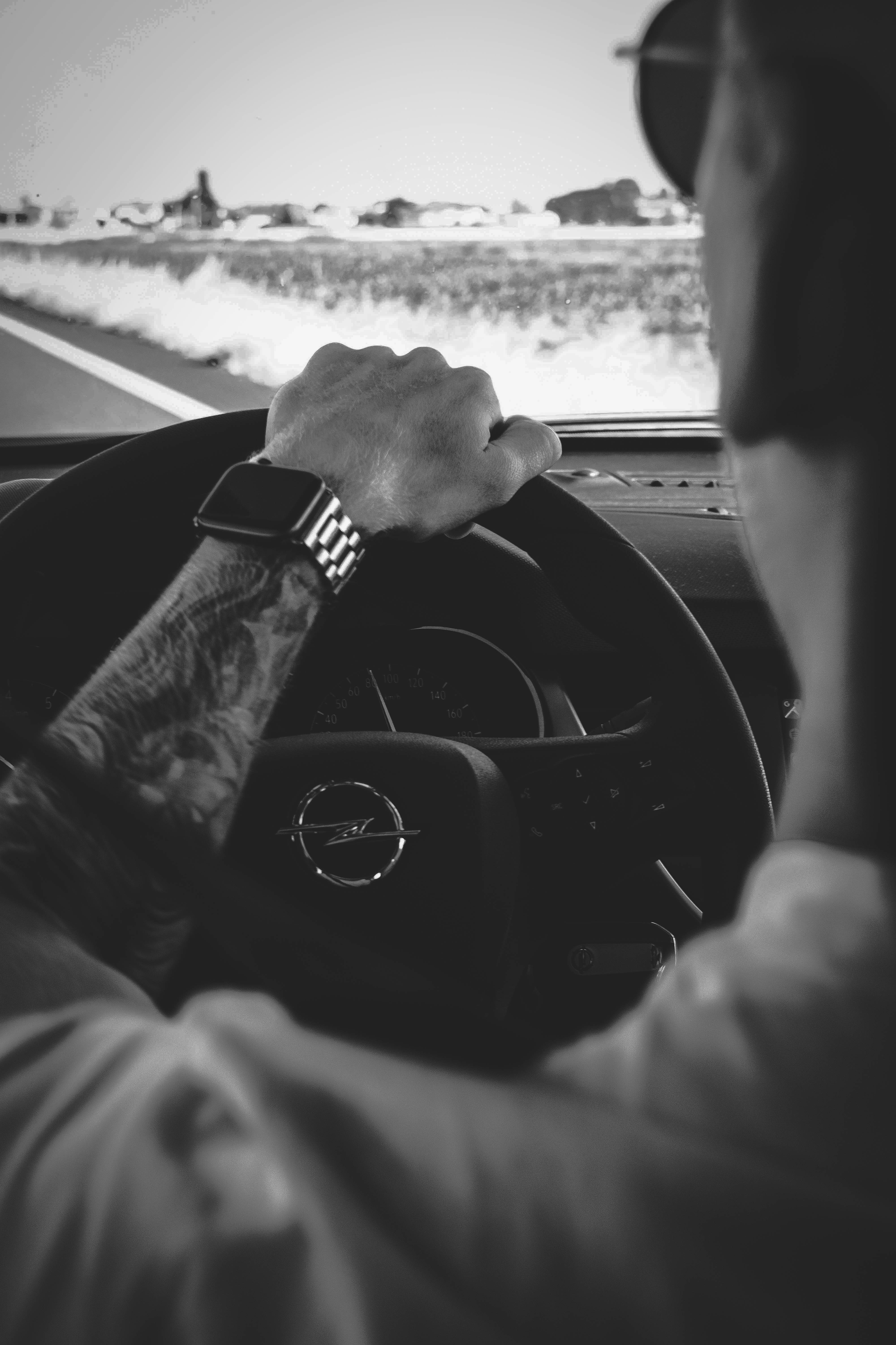 Close-up of a driver's hand gripping the steering wheel, showcasing intricate tattoos and a watch. The background features a blurred road, emphasizing movement.