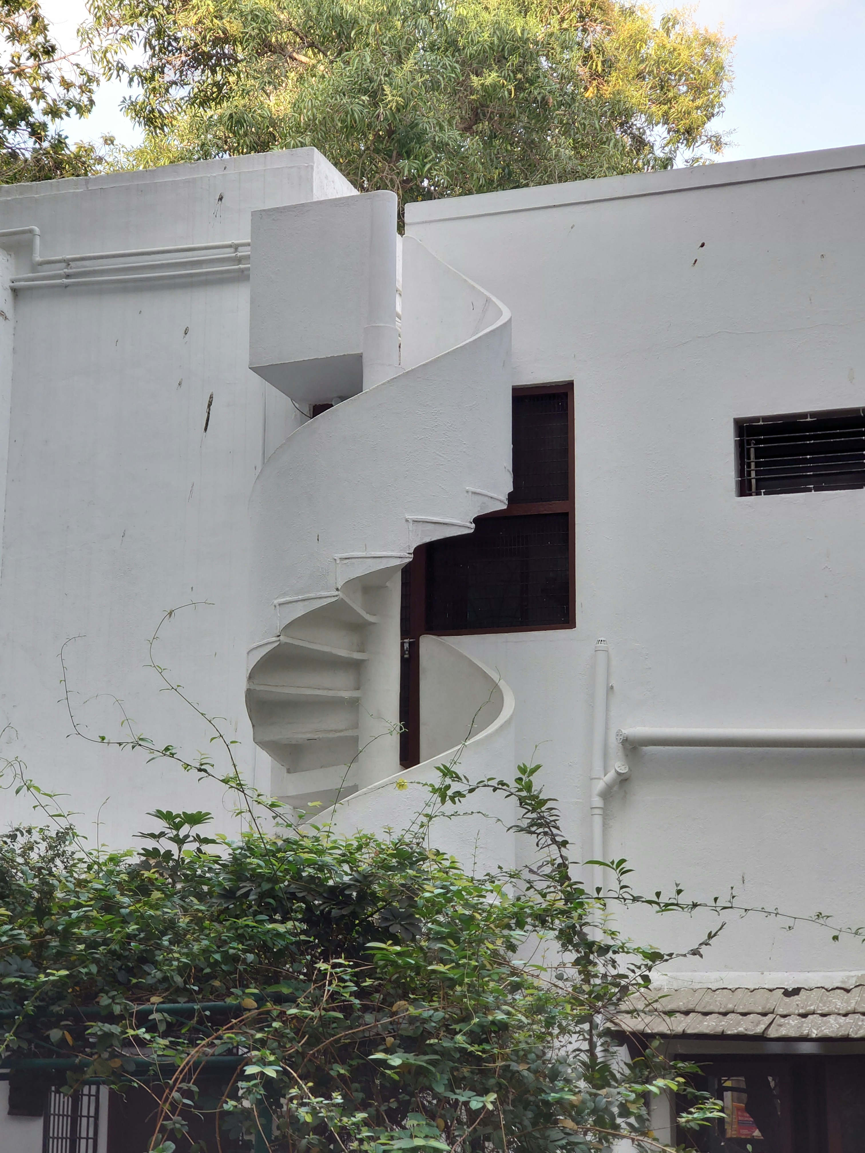 A spiral staircase gracefully ascends the side of a white building, framed by lush greenery below. The design highlights a blend of modern aesthetics and natural elements.