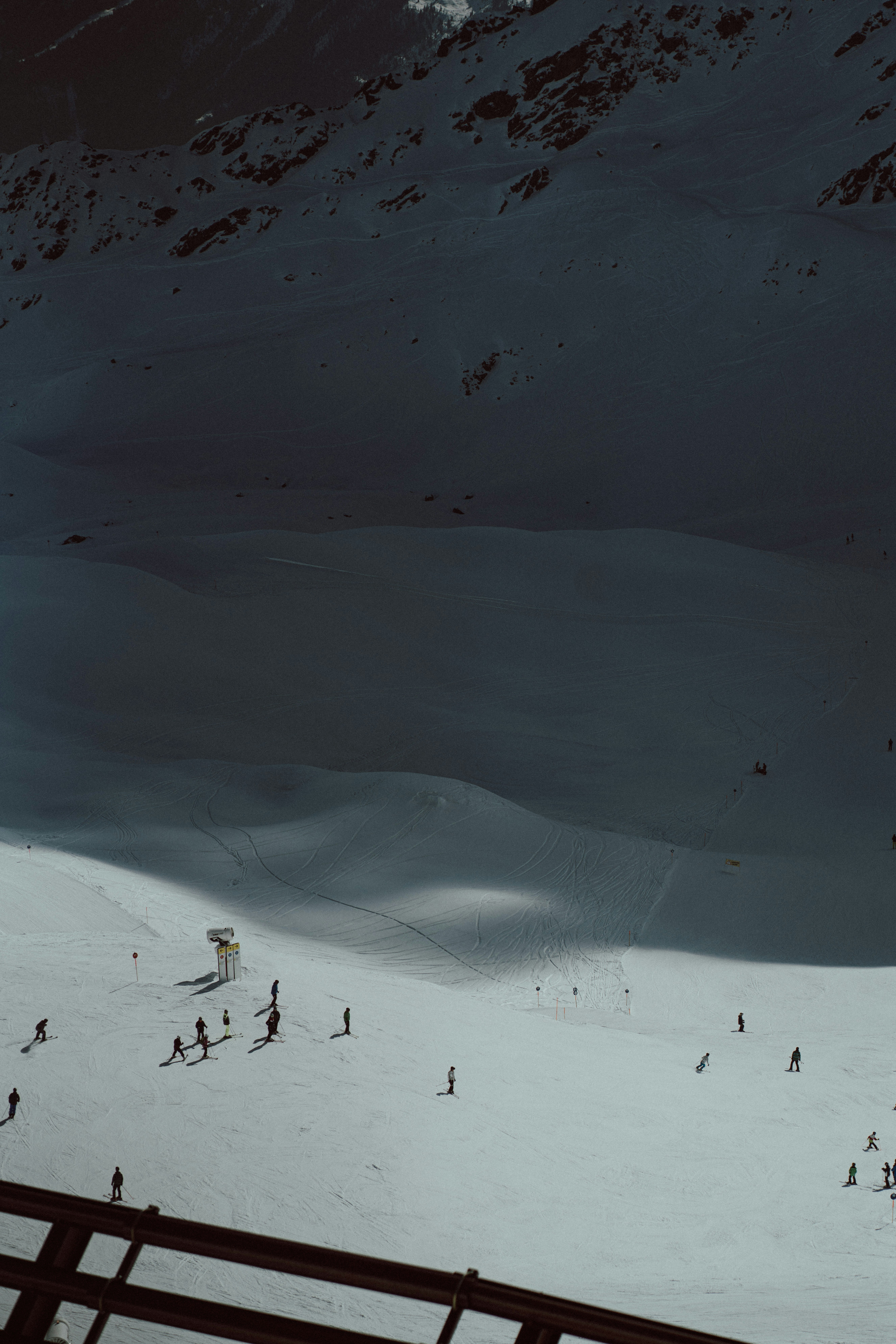 Skiers traverse a vast snowy landscape, with gentle slopes and shadows creating a serene atmosphere. A ski lift station stands in the foreground, hinting at the excitement of winter sports.