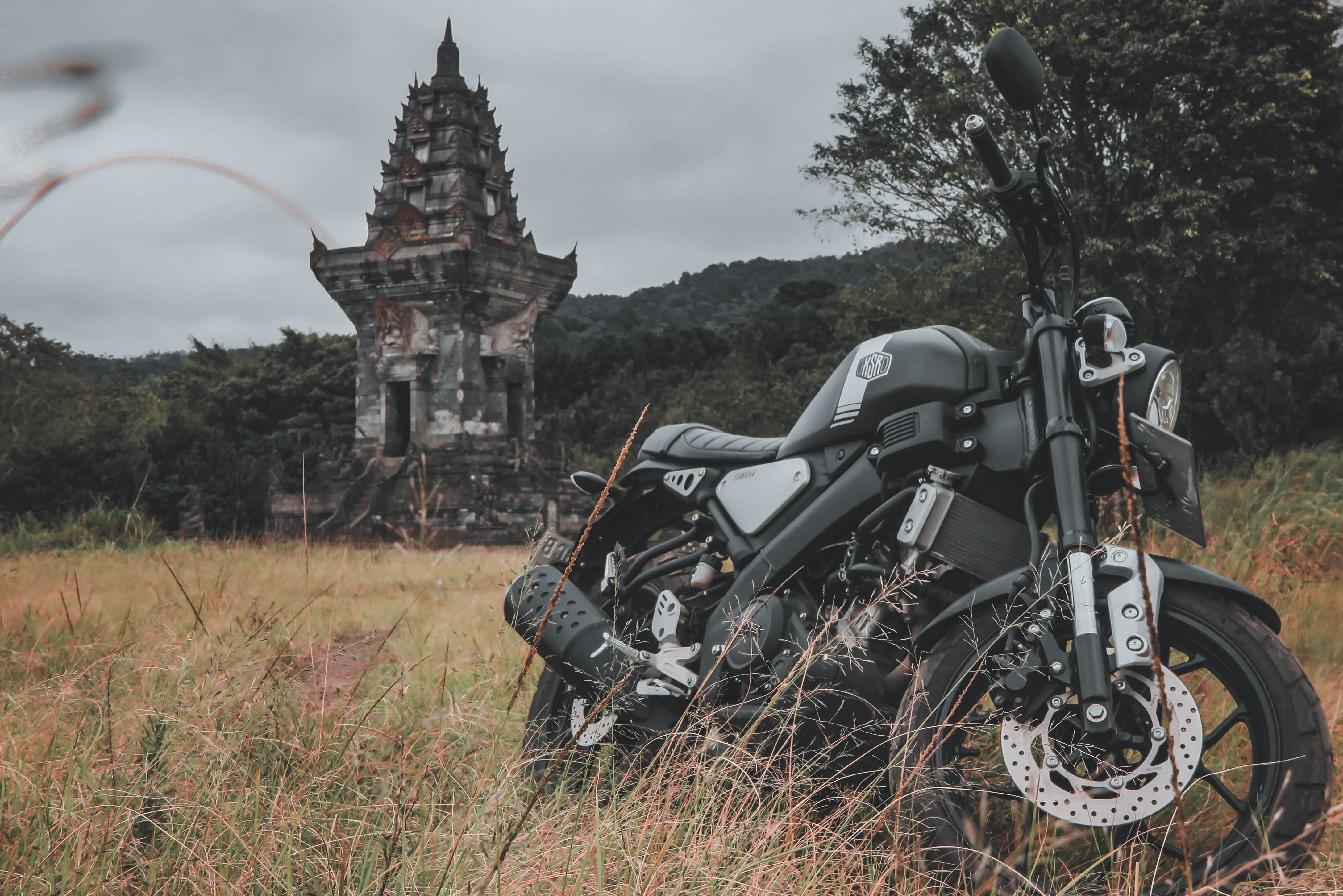 A sleek black motorcycle stands in tall grass, with a historic temple in the background under a cloudy sky.