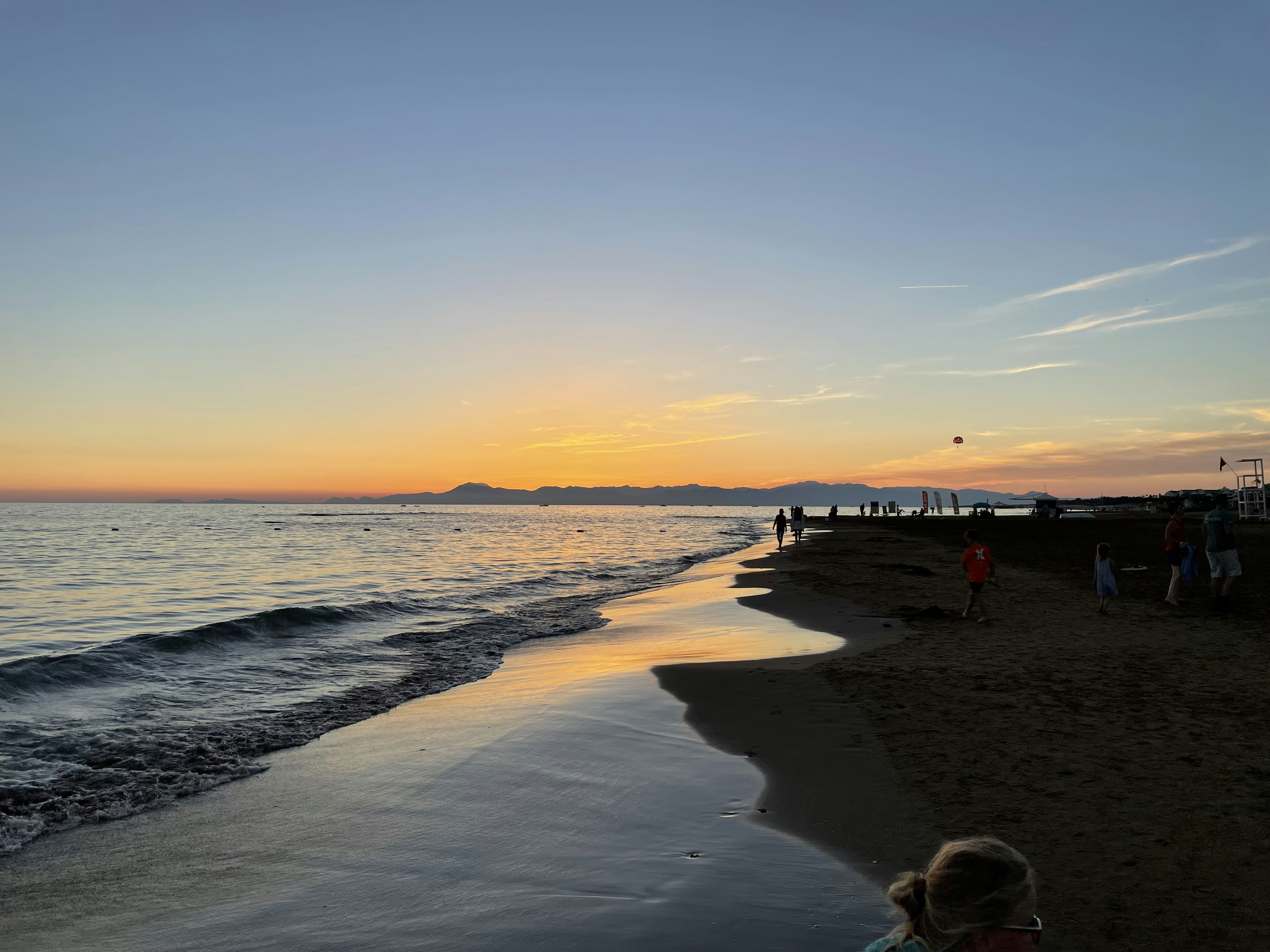 Sunset over a tranquil beach with gentle waves and silhouettes of distant hills.
