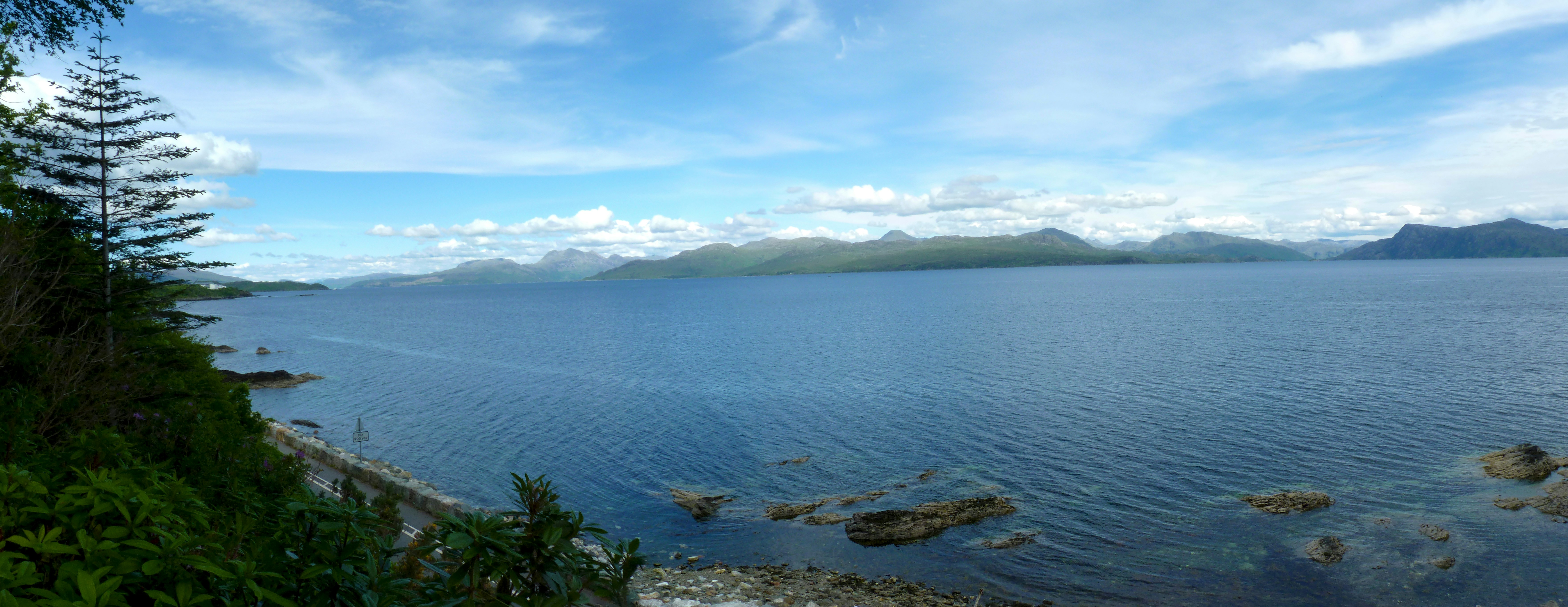 Expansive view of a calm blue sea bordered by a lush green coastline and distant mountain range under a bright sky.