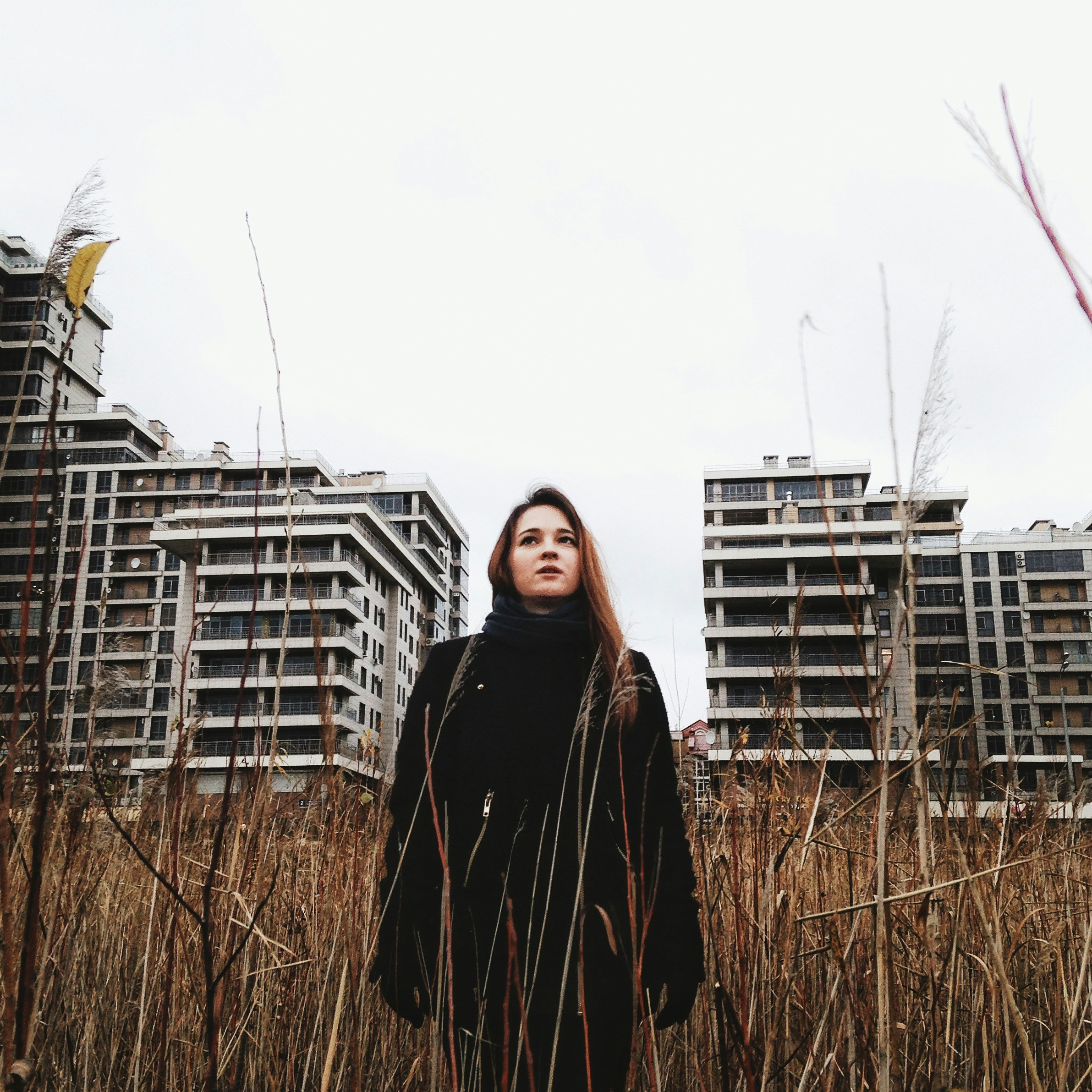 Shelter Amid Towers: A Woman in Tall Grass Beneath a Grey Sky