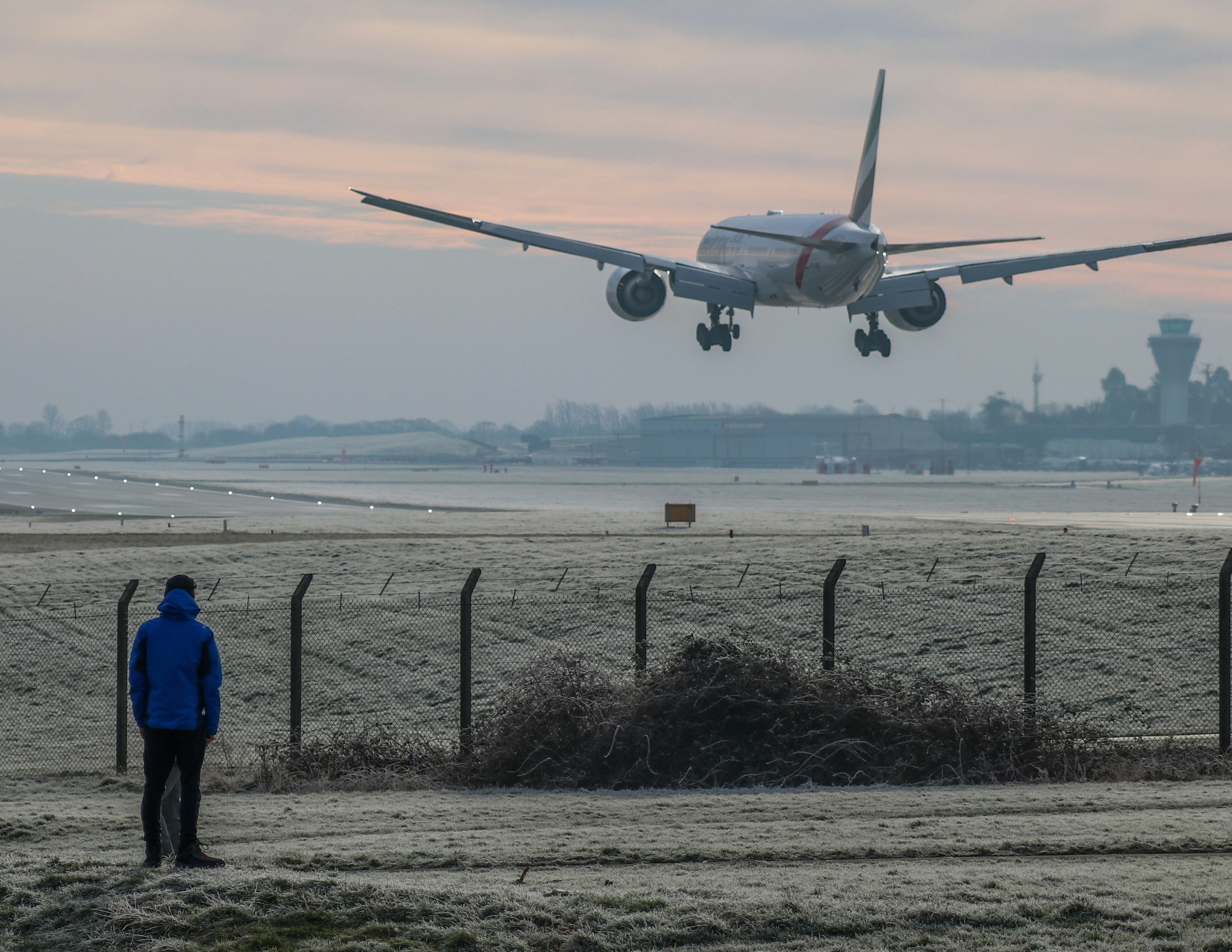 Person in blue jacket watches airplane descending towards runway on a frosty morning.