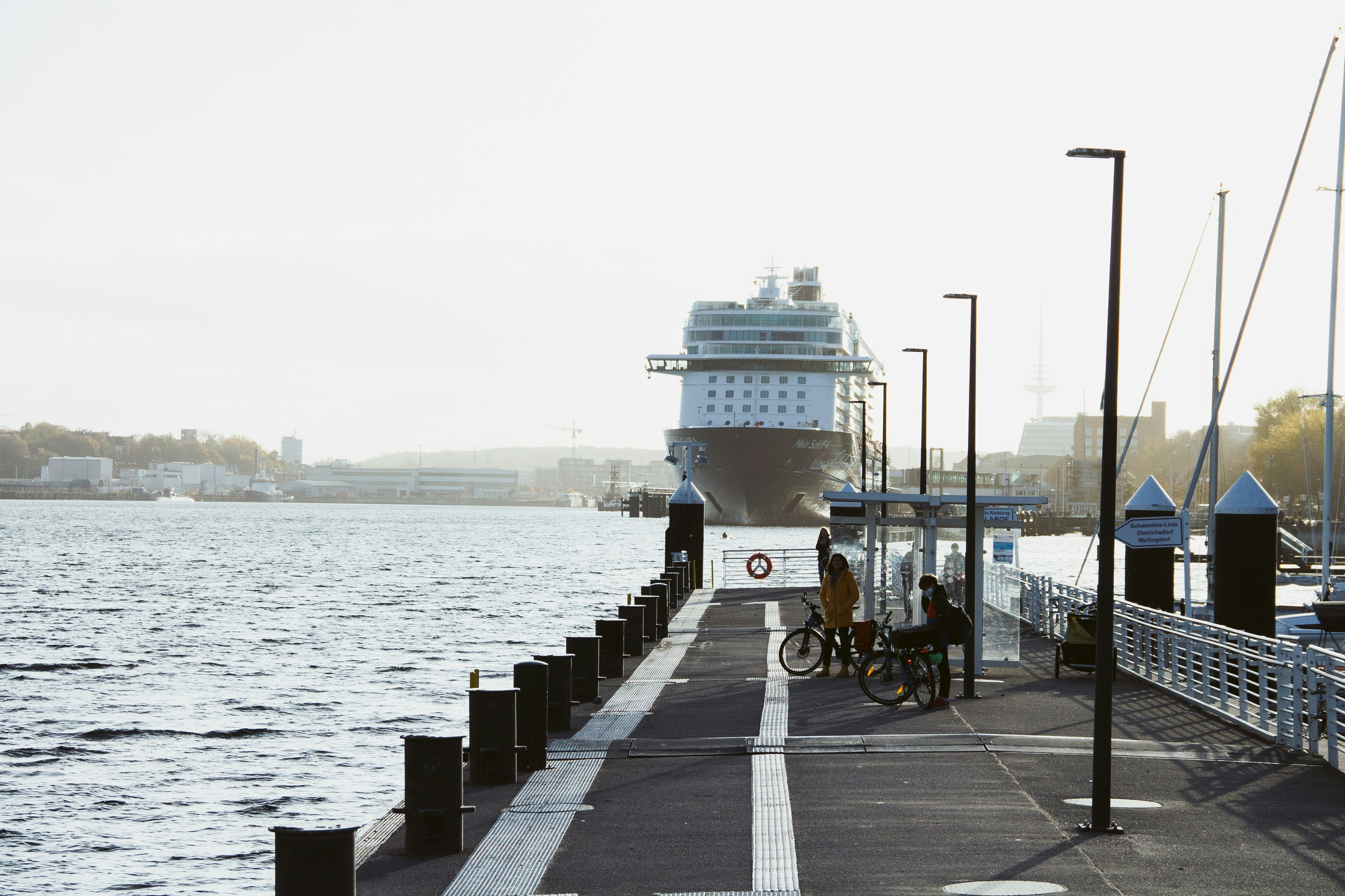 Cruise ship docked at a pier under a bright sky with calm waters and distant shoreline.