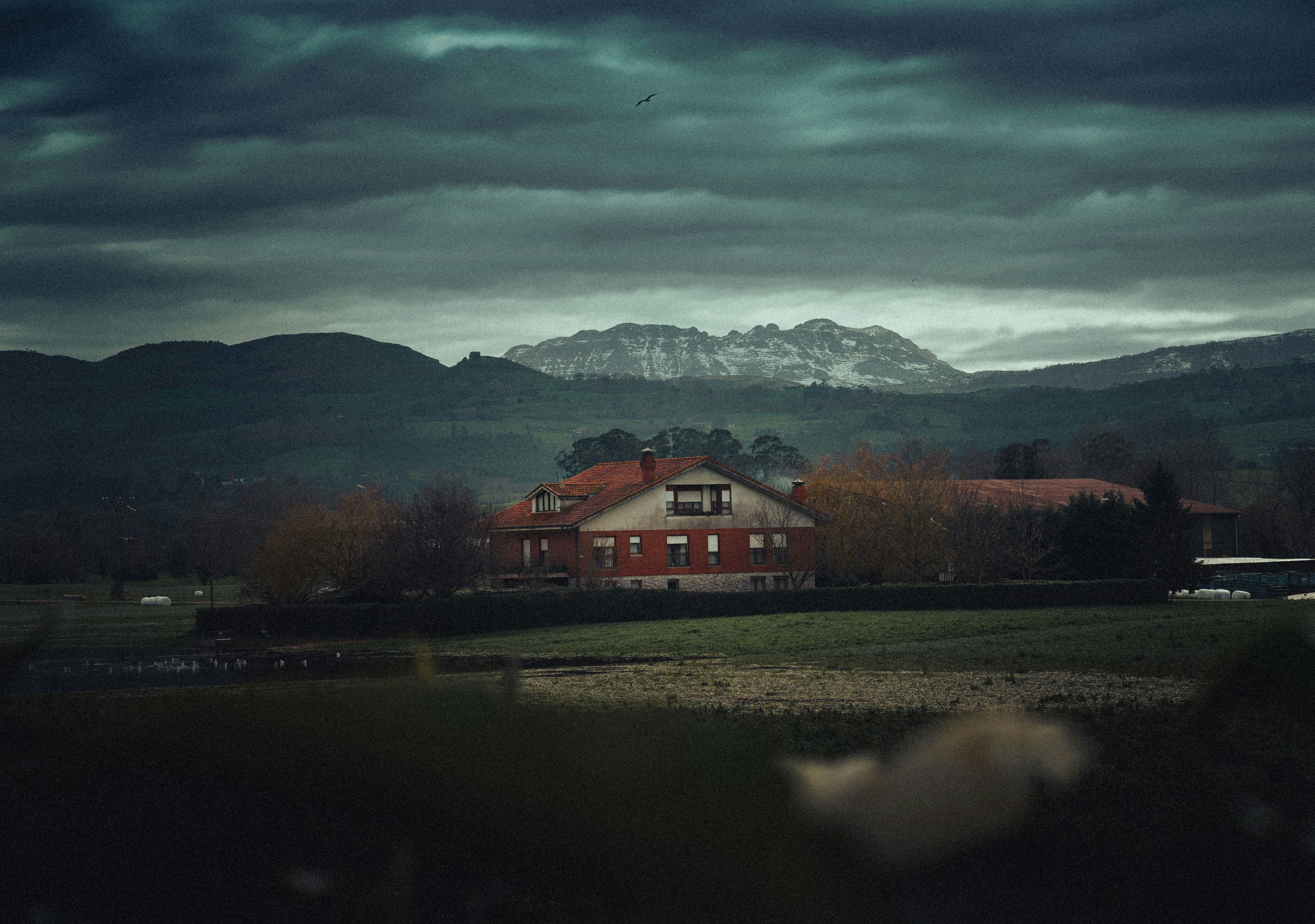 A rustic red house nestled in a valley, framed by looming mountains and dark, moody skies. Autumn foliage hints at the changing seasons.