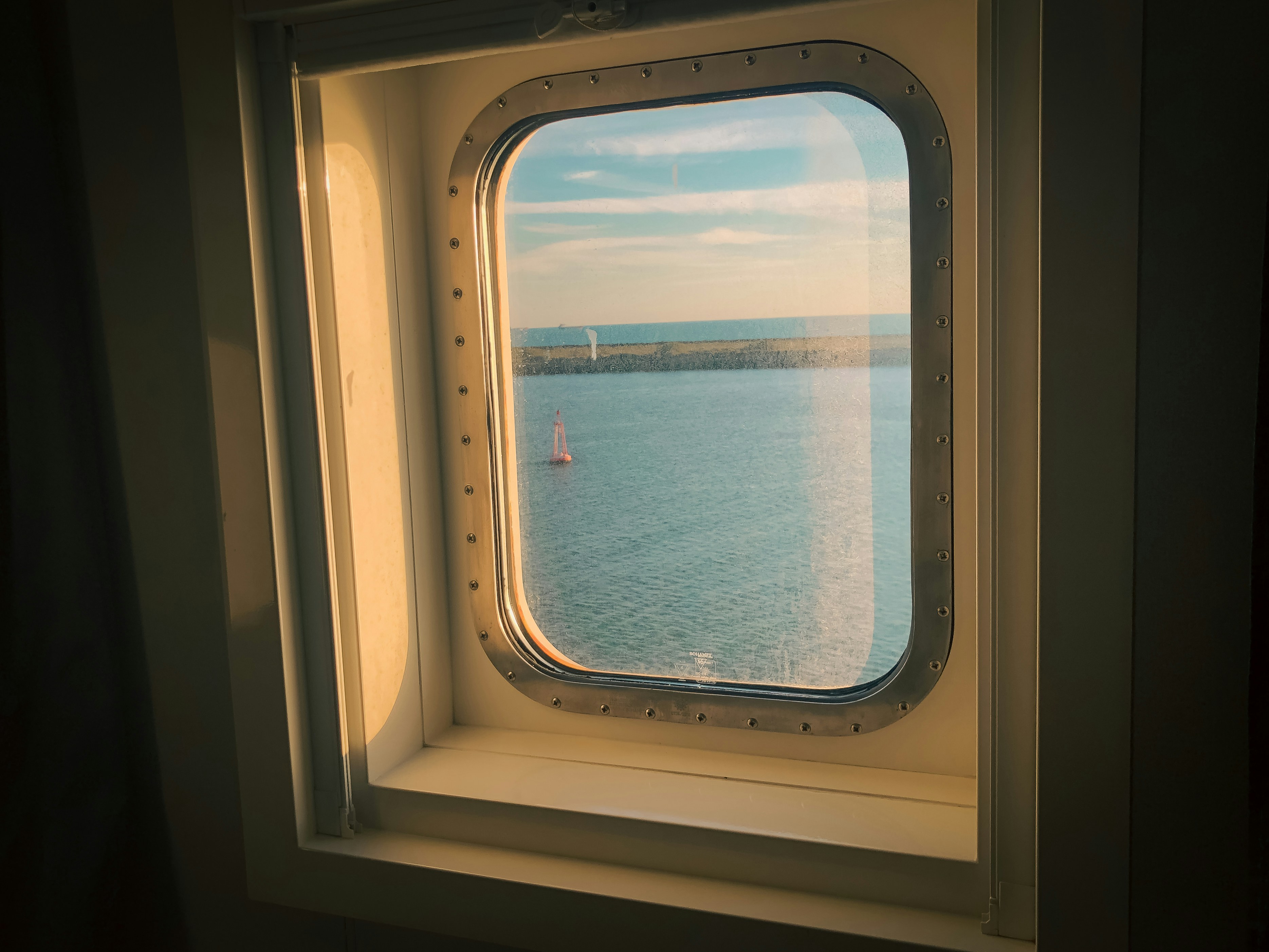View through a ship's porthole of calm waters and distant shoreline under a pastel sky.