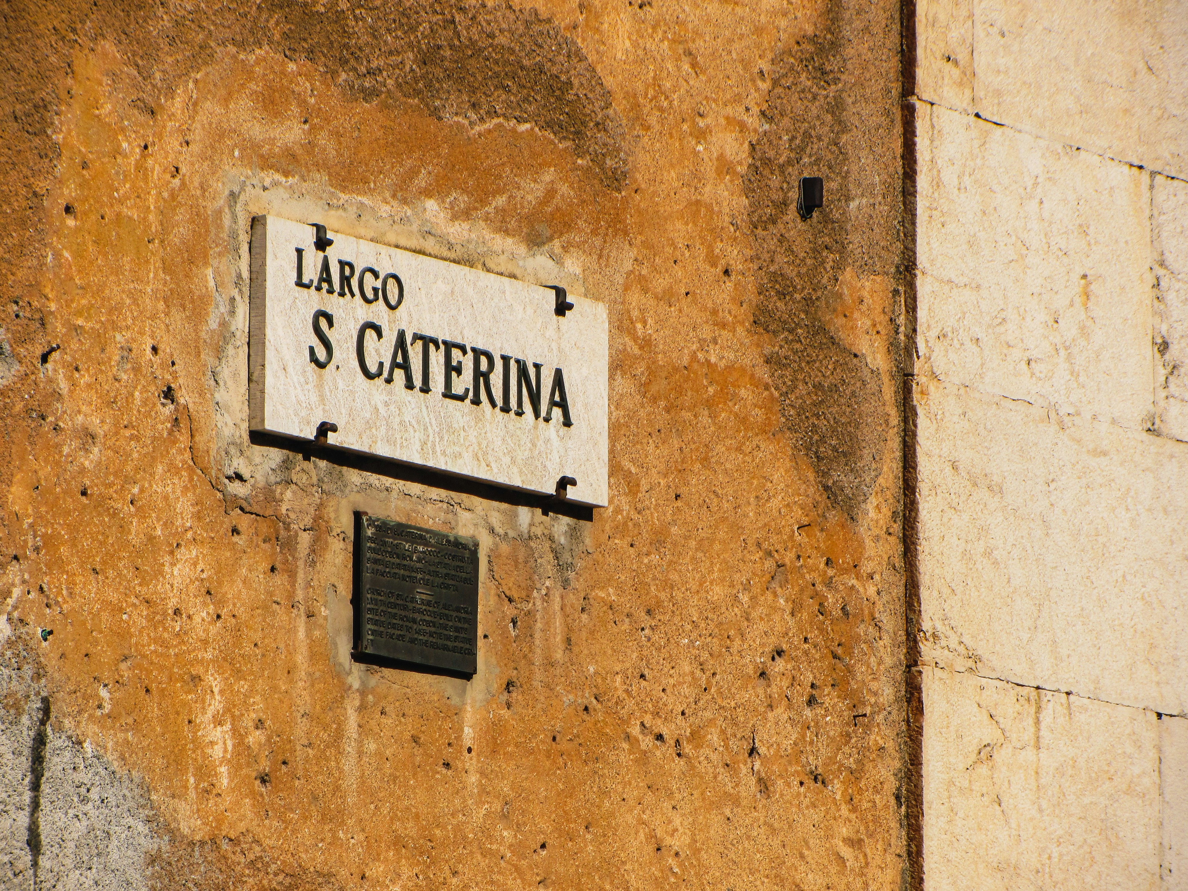 Weathered street sign mounted on a textured, ancient wall in warm sunlight.