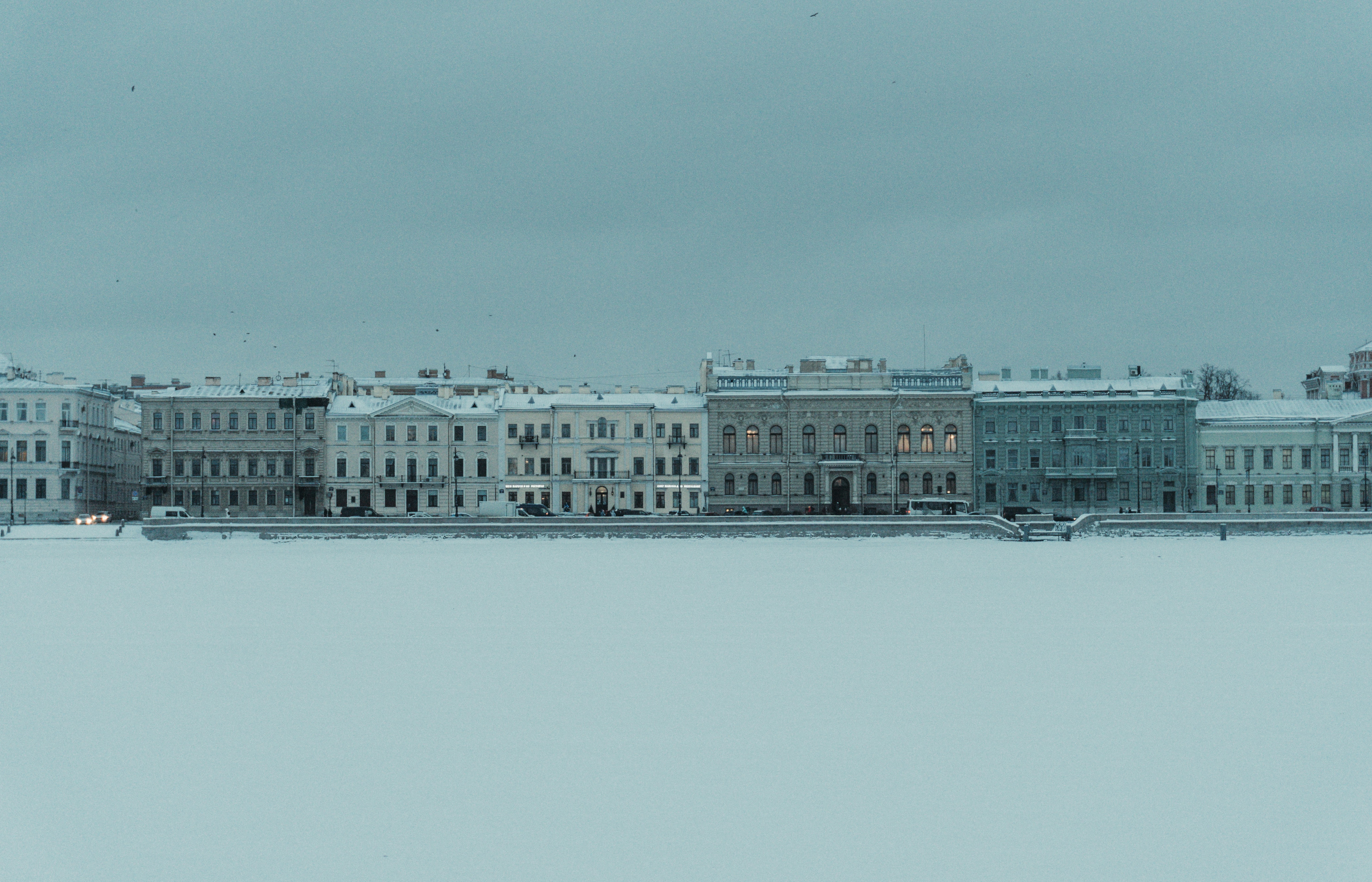 A winter cityscape photograph of a row of neoclassical buildings along a snow-covered river under a pale blue sky. The scene emphasizes calm, cool tones and a clean horizon.