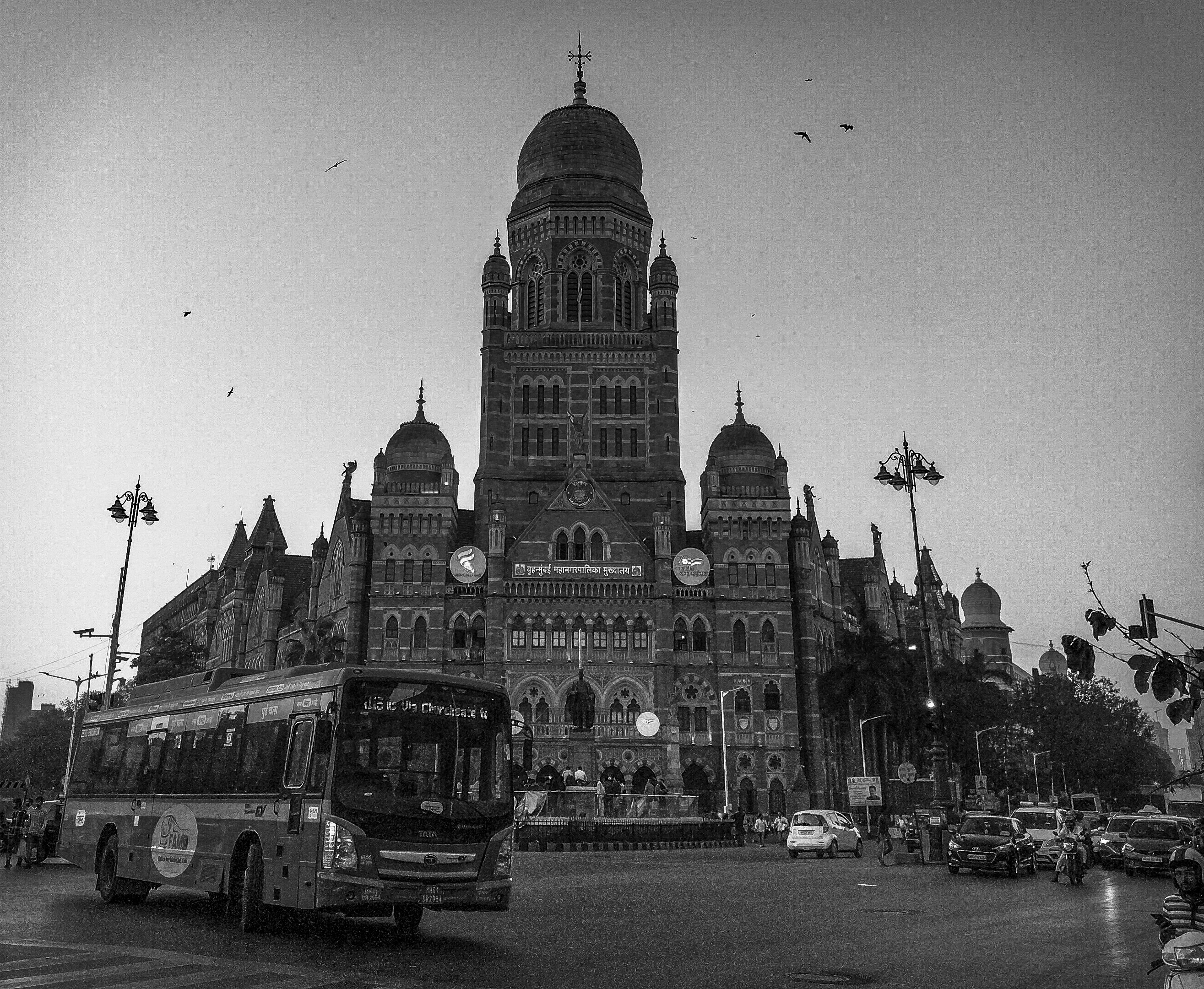 Majestic Gothic-style building with bustling traffic and a city bus in the foreground, captured in monochrome.