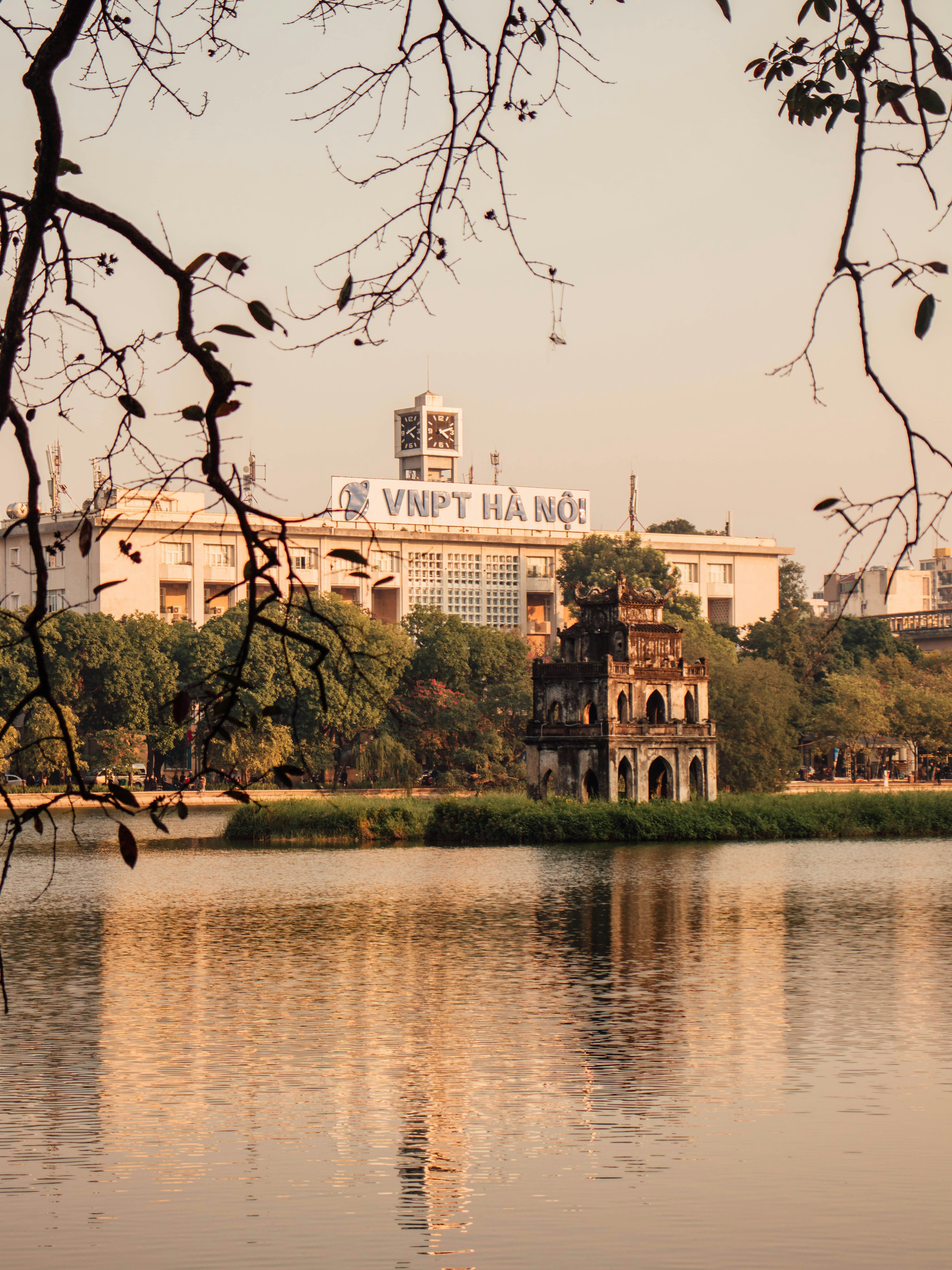 Historic tower reflected in the tranquil waters, framed by branches, with modern architecture in the background. 