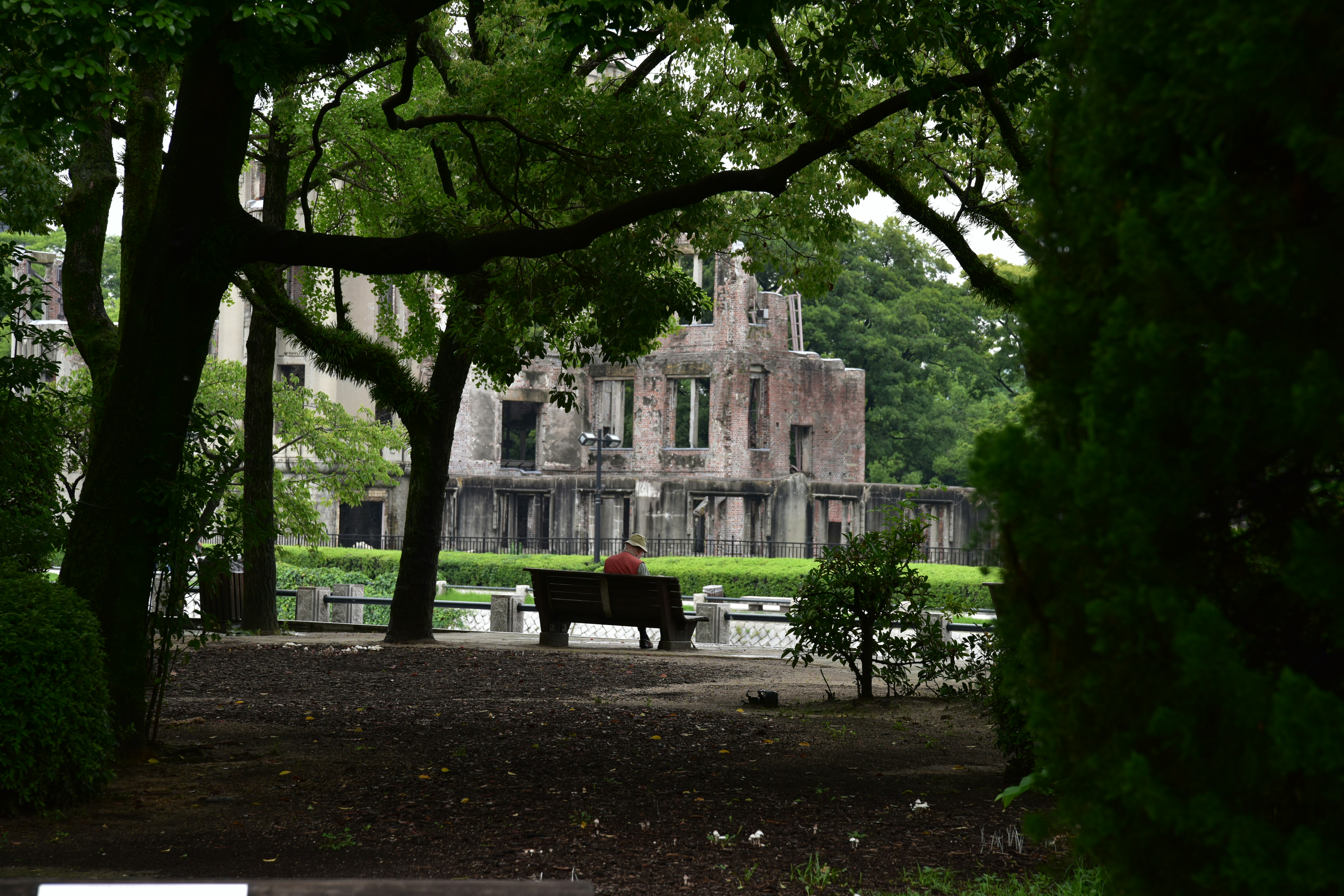A lone figure sits on a bench under lush trees near historic building ruins, framed by greenery.