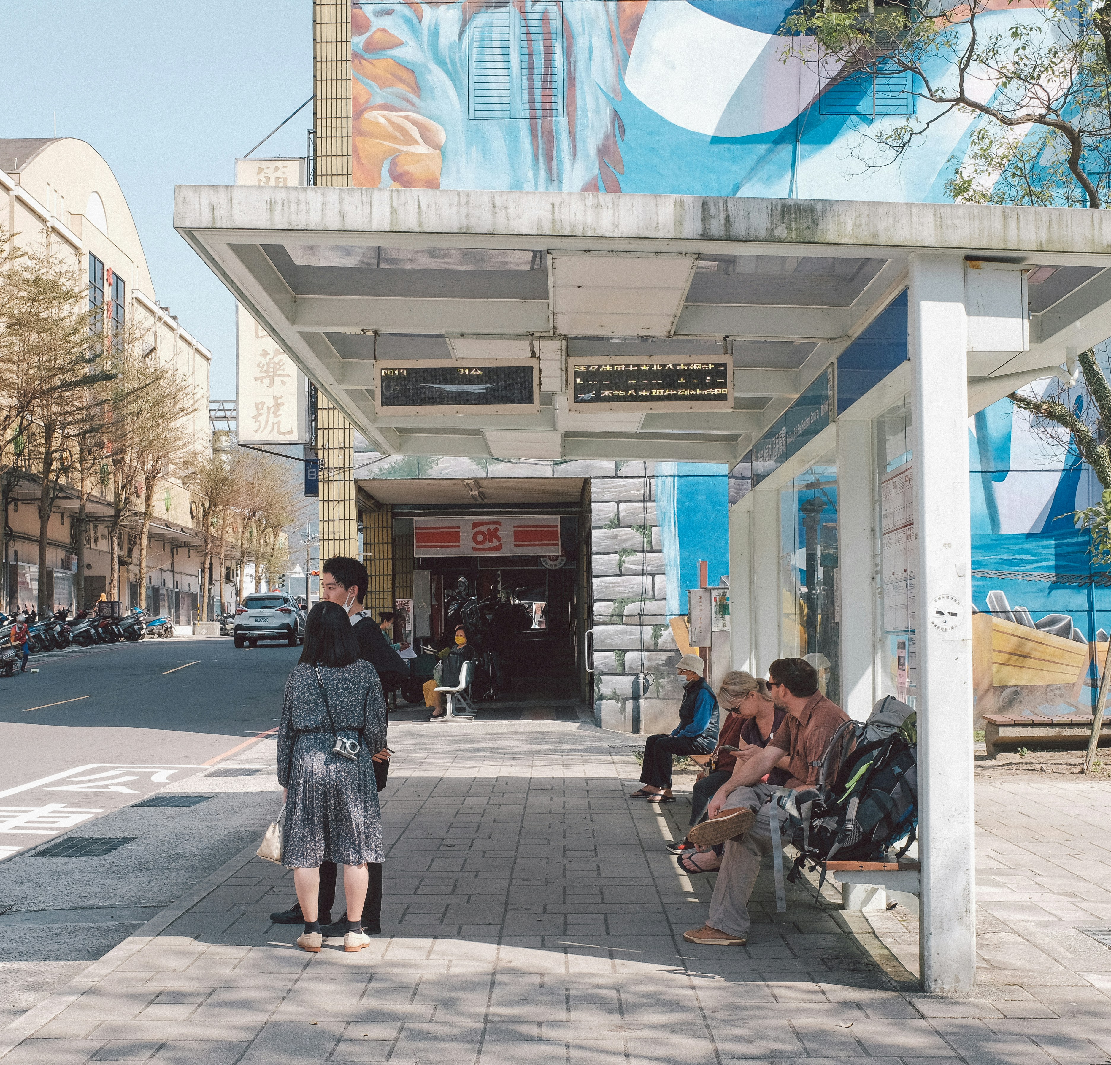 A vibrant mural adorns a bus stop while commuters engage in conversation, showcasing daily life in a bustling urban setting.