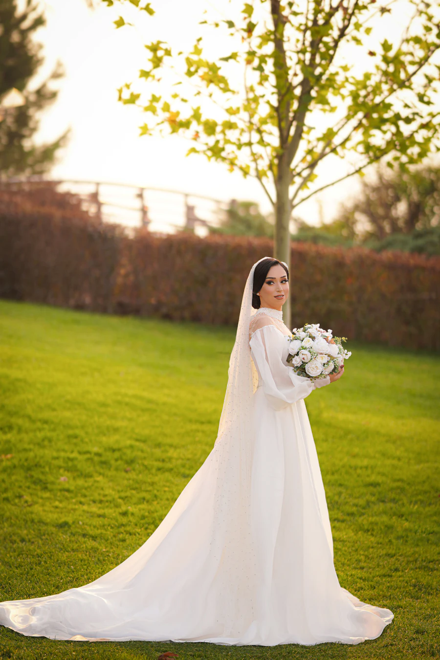 A bride in a flowing white gown and veil holds a bouquet on a bright green lawn, with warm sunset light and a tree behind her.