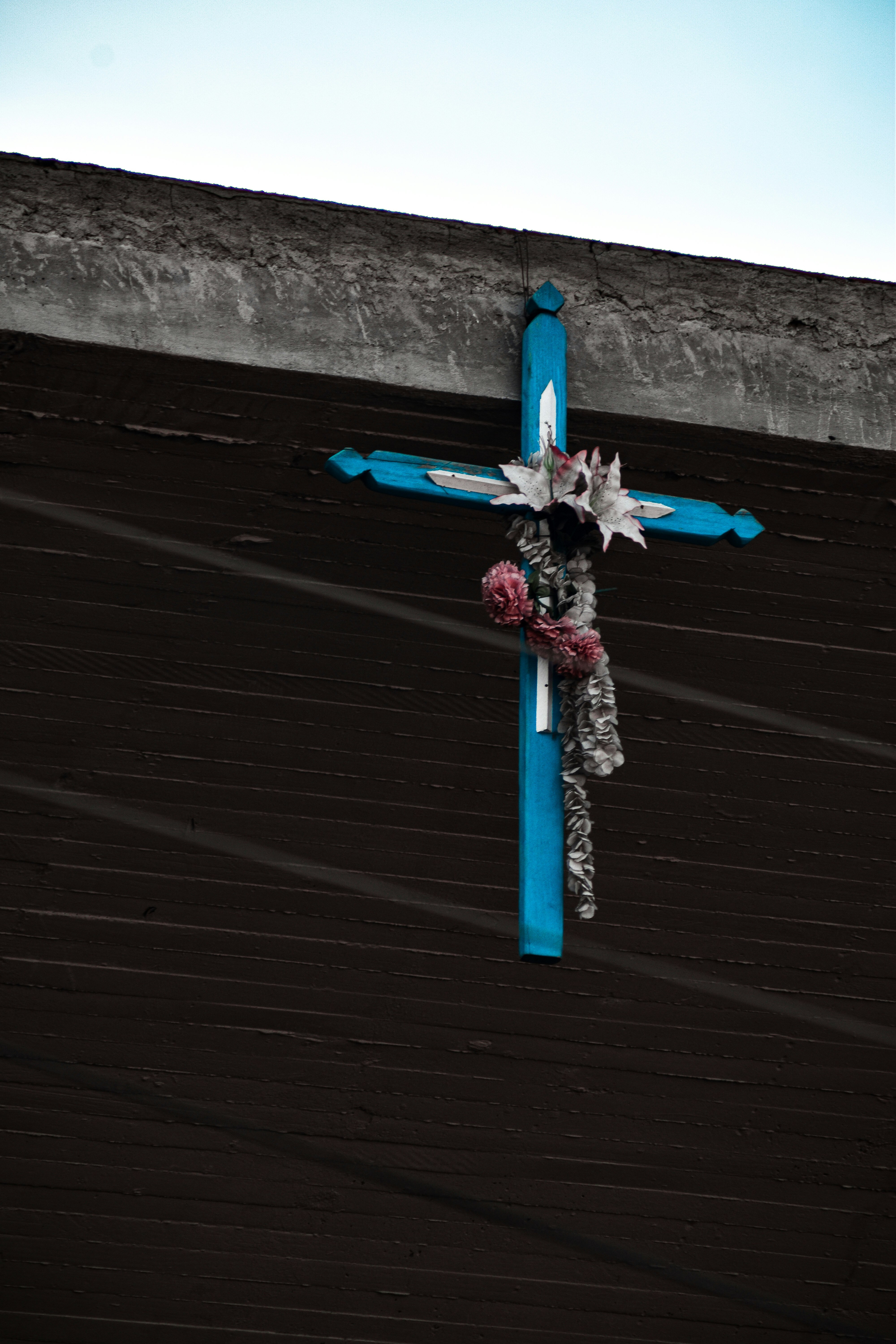 Blue crucifix mounted on weathered dark wood, adorned with dried flowers and fabric, against a pale sky.
