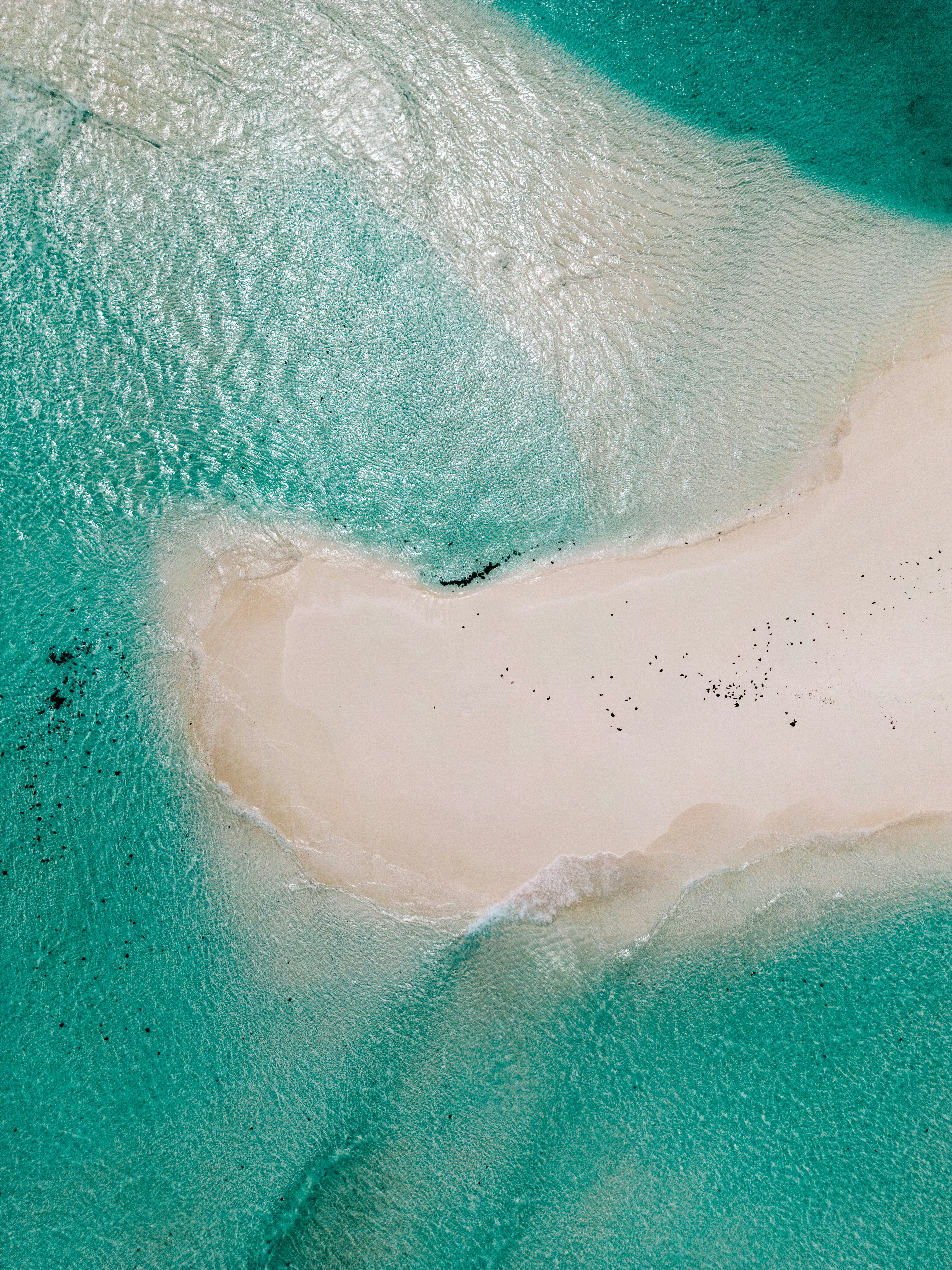 Aerial drone photograph showing a pale sandbar curving through turquoise shallow water, with rippling surf along its edge.