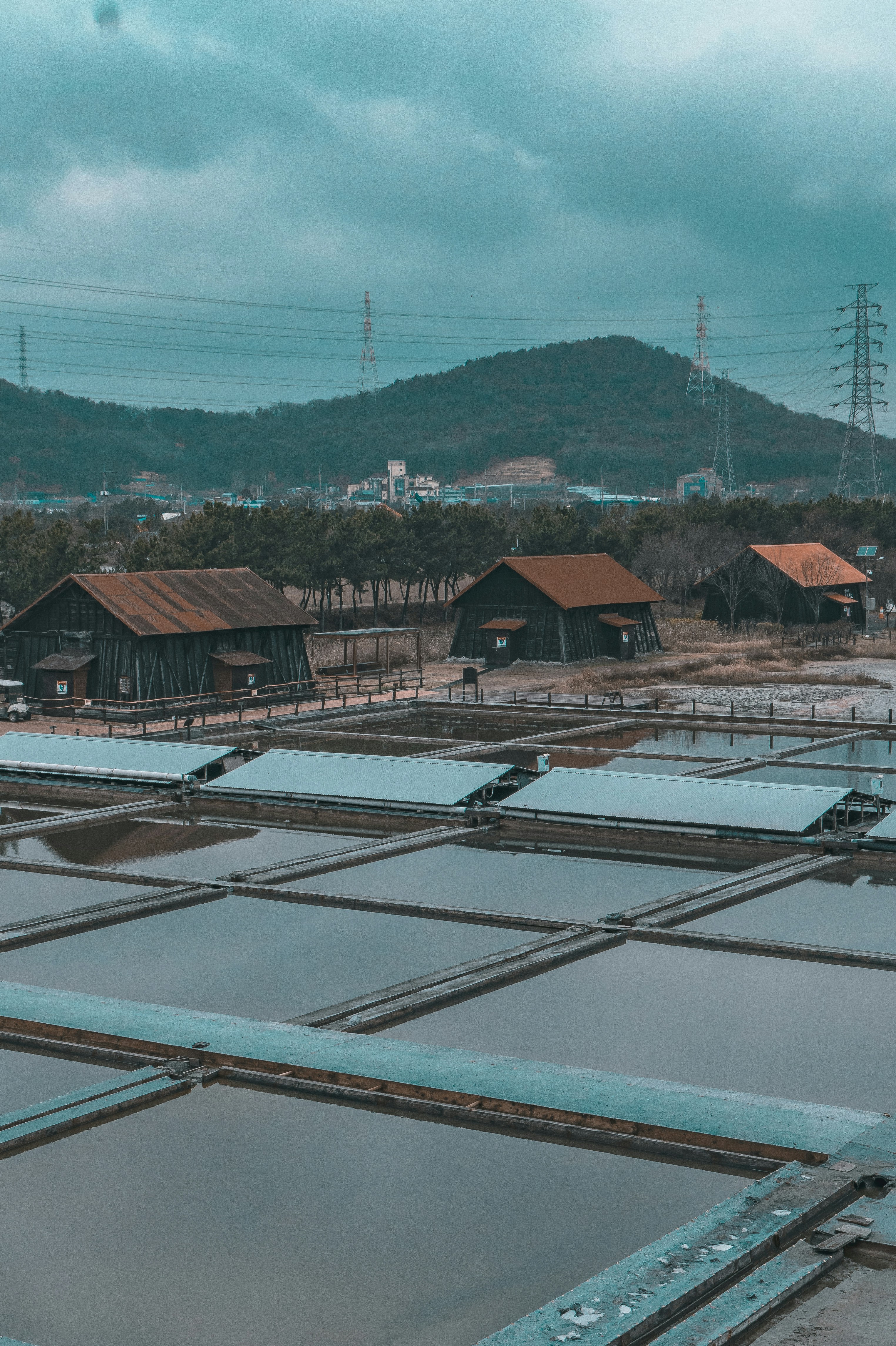 Rustic wooden huts surround expansive salt flats, reflecting the overcast sky. Power lines stretch across the landscape, juxtaposing nature and industry.