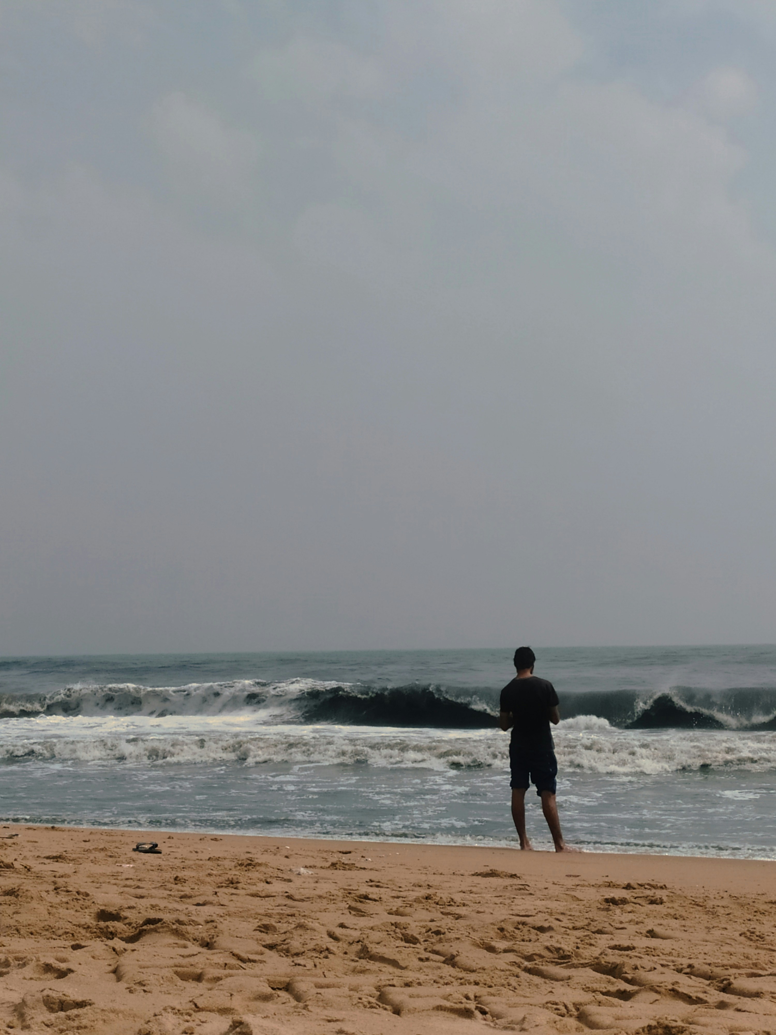 A solitary figure stands on the beach, gazing at the crashing waves under a hazy sky.