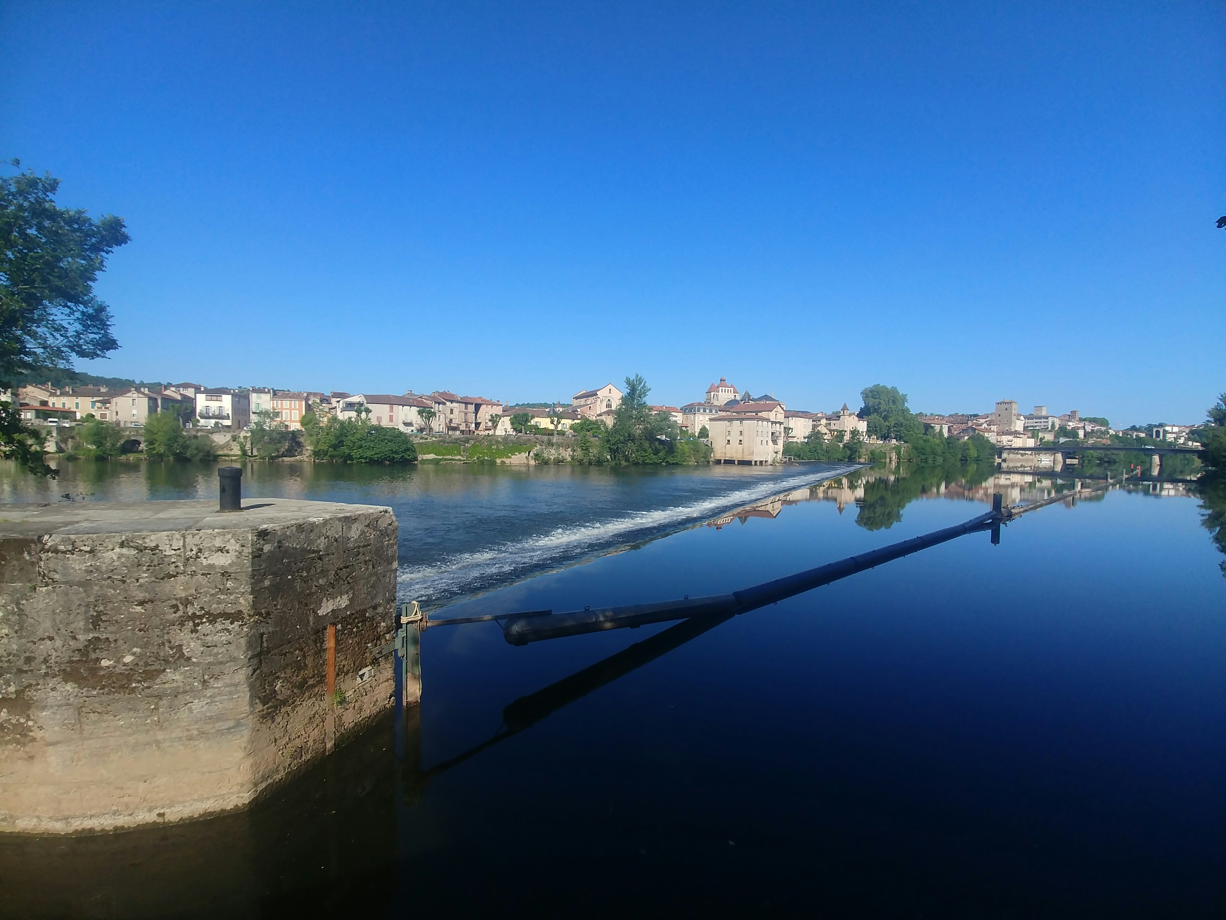 Calm river reflecting blue sky and historic buildings under bright sunlight.