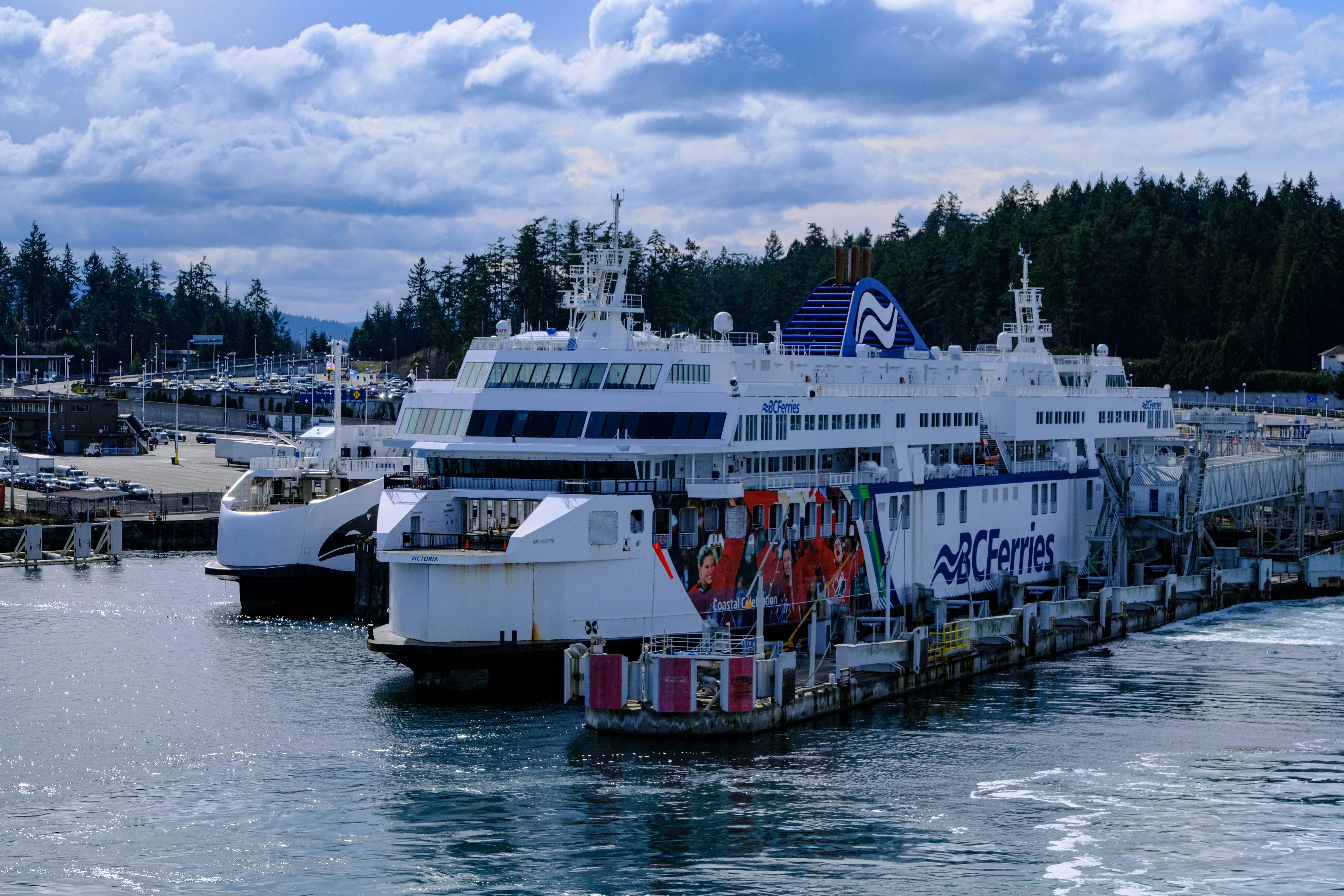 BC Ferries vessel docked at a busy terminal, surrounded by other ferries and a scenic backdrop of trees and clouds.