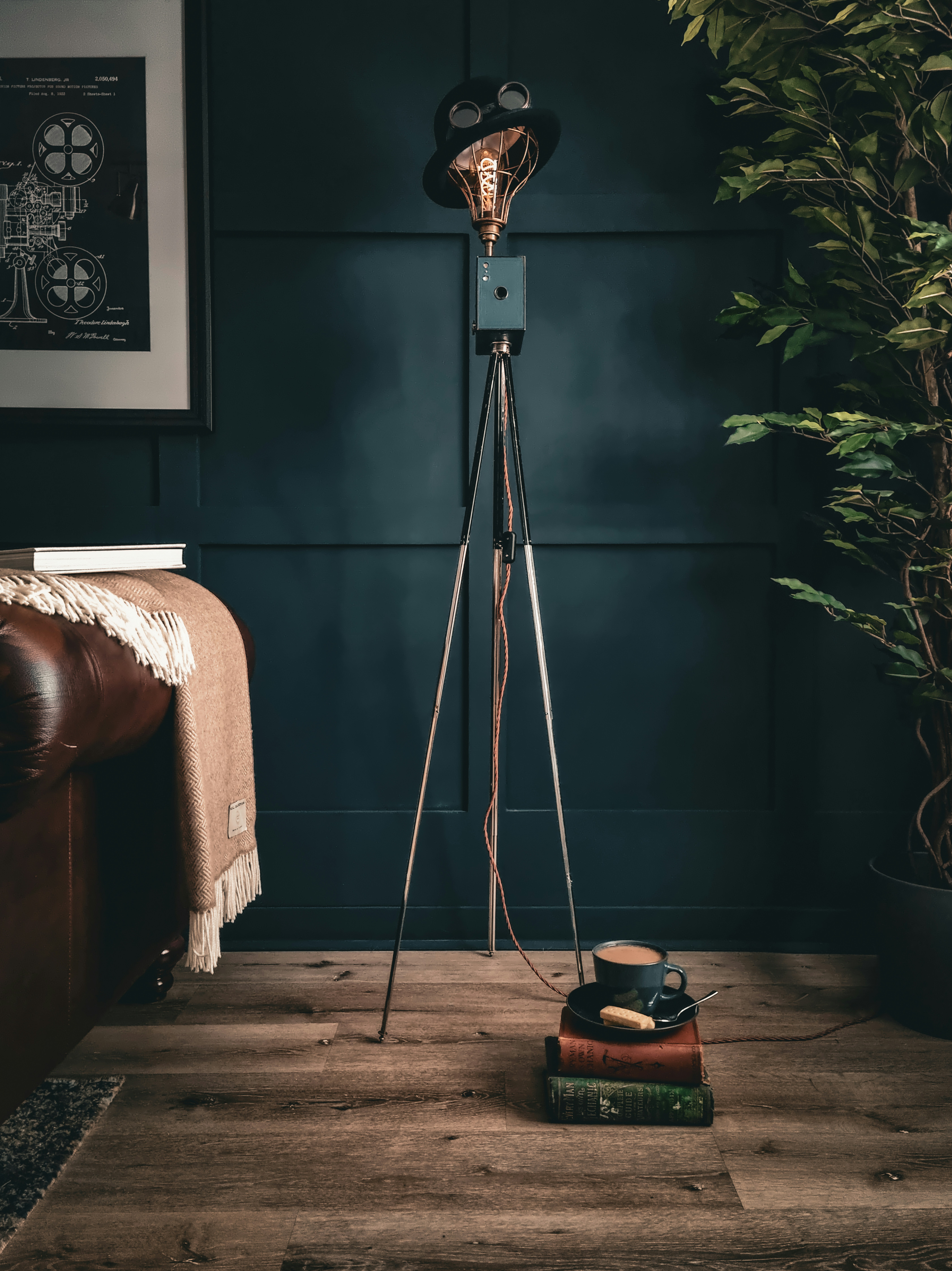 A stylish floor lamp casts warm light in a cozy living space, complemented by a stack of books and a mug on a table. The deep blue wall adds a touch of sophistication.