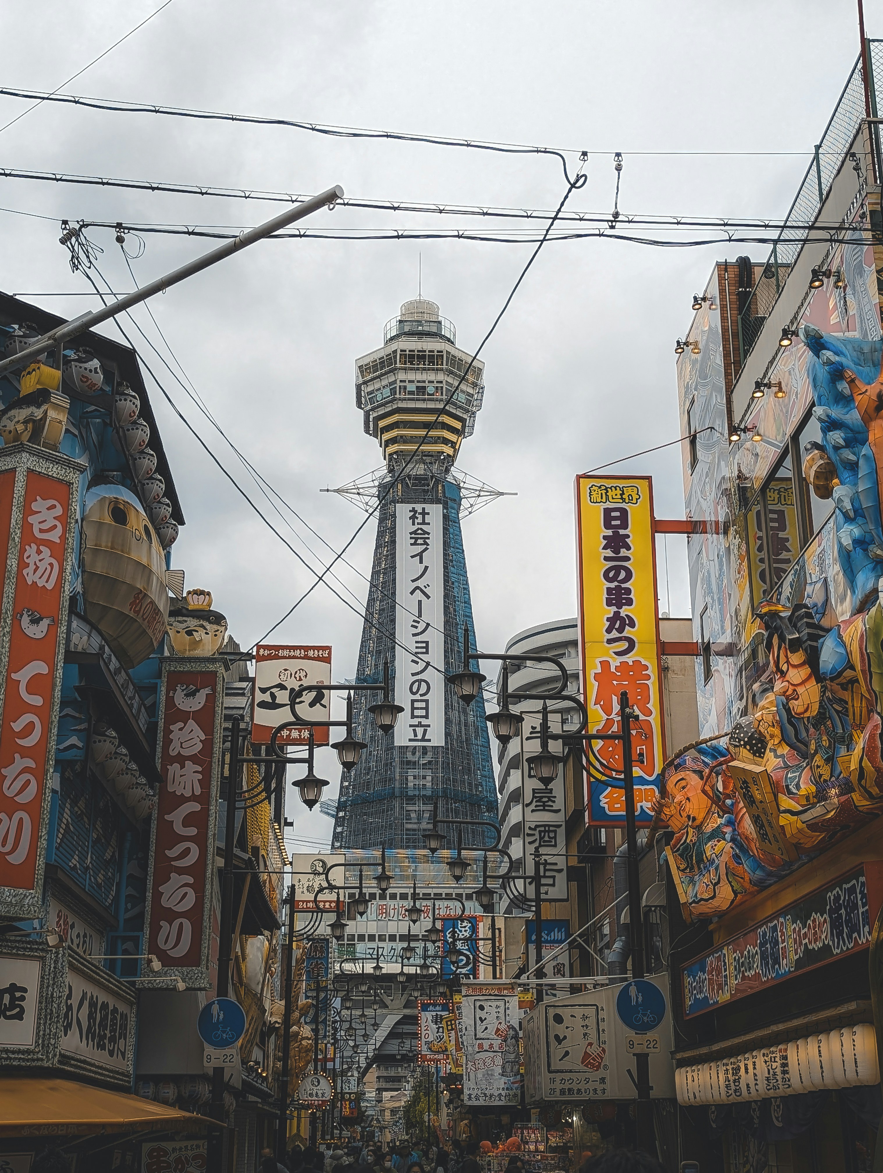 Photograph of a bustling Shinsekai street in Osaka, with vibrant signs along both sides and the Tsutenkaku Tower rising at the center.