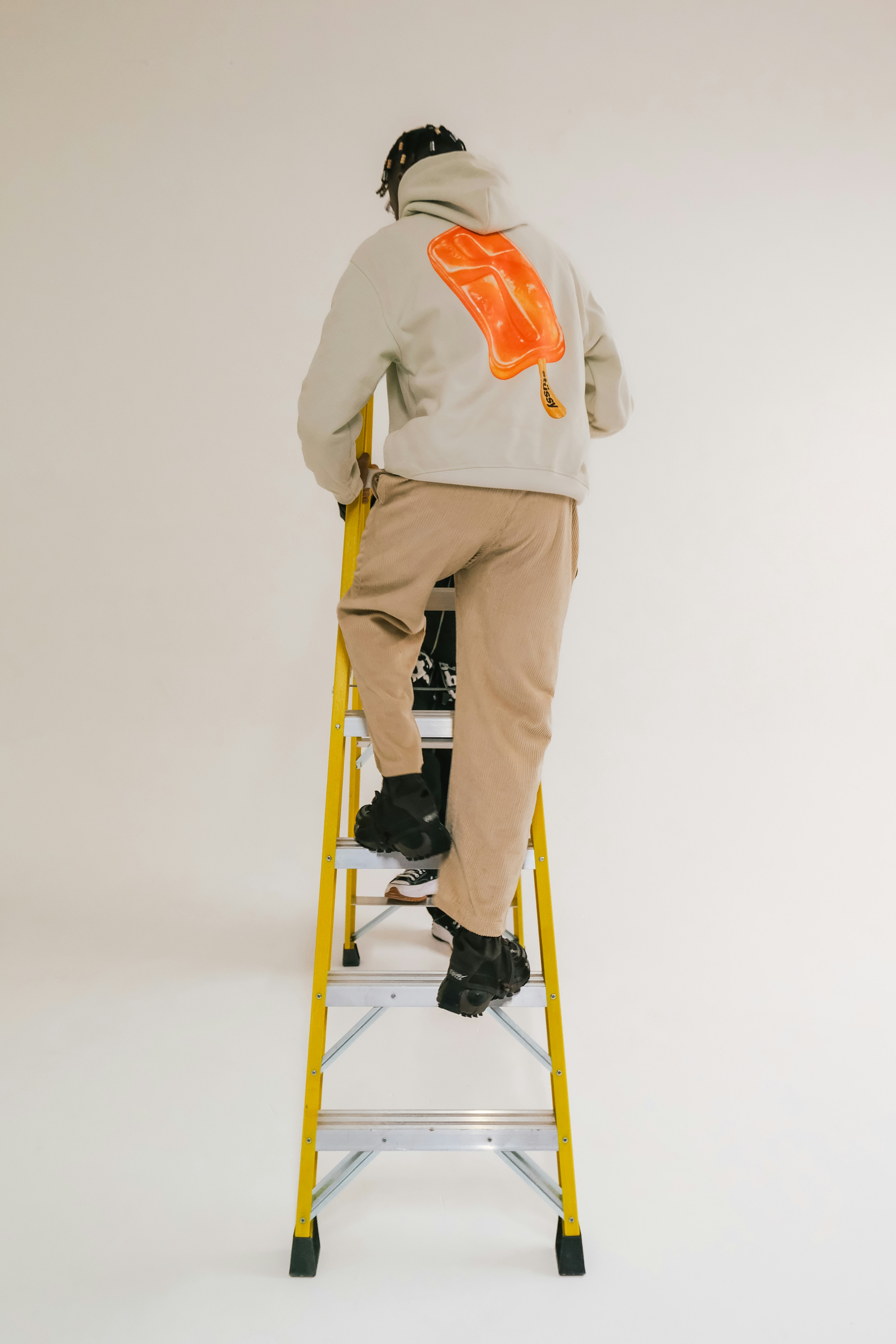Person in a hoodie with a popsicle graphic stands on a yellow ladder against a white backdrop.