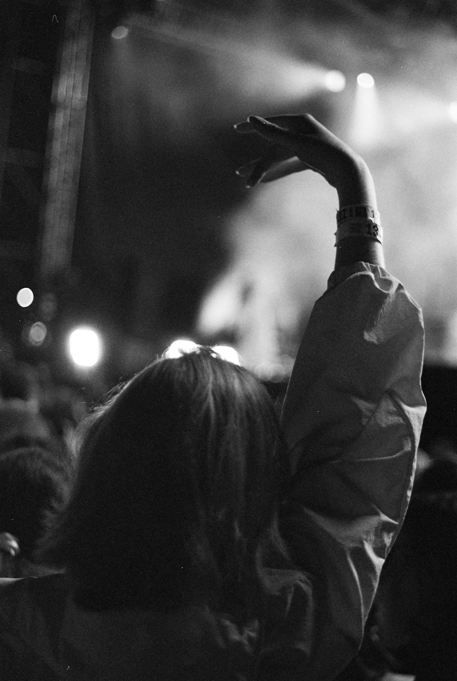 Backlit concertgoer with an arm raised overhead amid haze and stage lights, seen from behind in monochrome. Blurred crowd and bright beams create a moody live-show atmosphere.