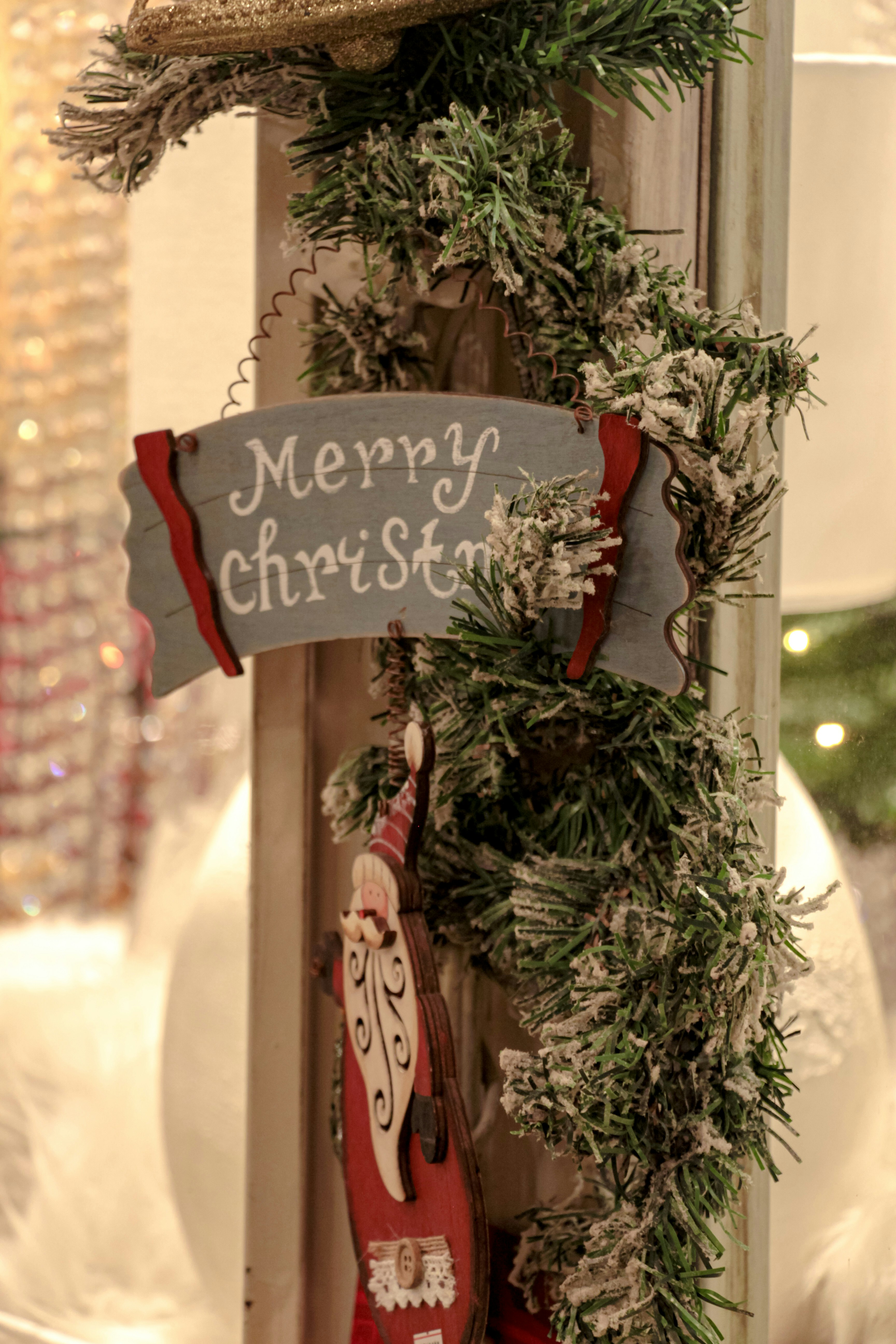 A frosted evergreen wreath frames a curved 'Merry Christmas' sign with a hanging Santa ornament. The festive display sits against a softly blurred background.