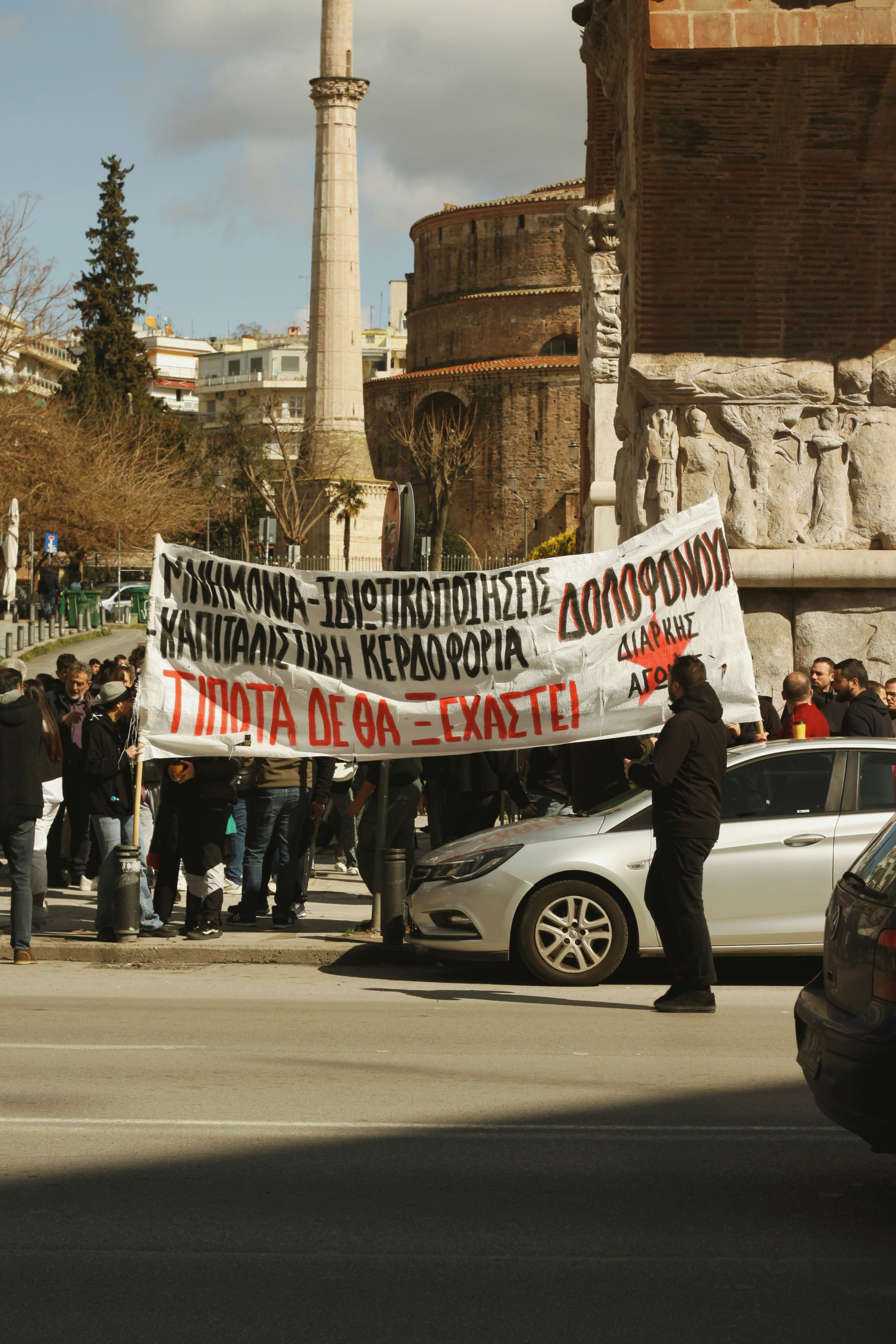 Bannered protest along a sunlit Athens avenue near ancient pillars.