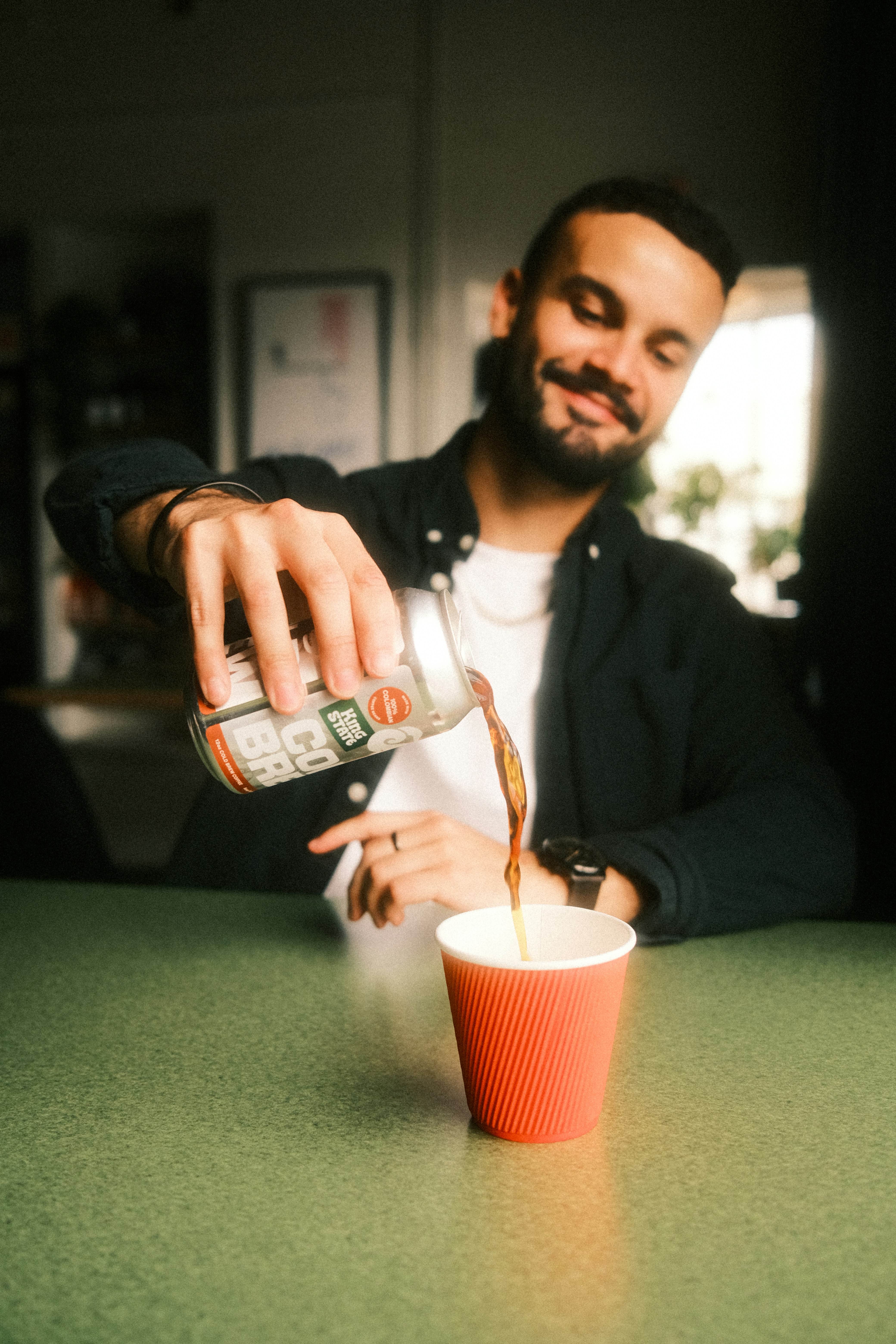 A man pours a dark beverage from a can into a red paper cup, showcasing the fluid motion and focus on the act of serving.