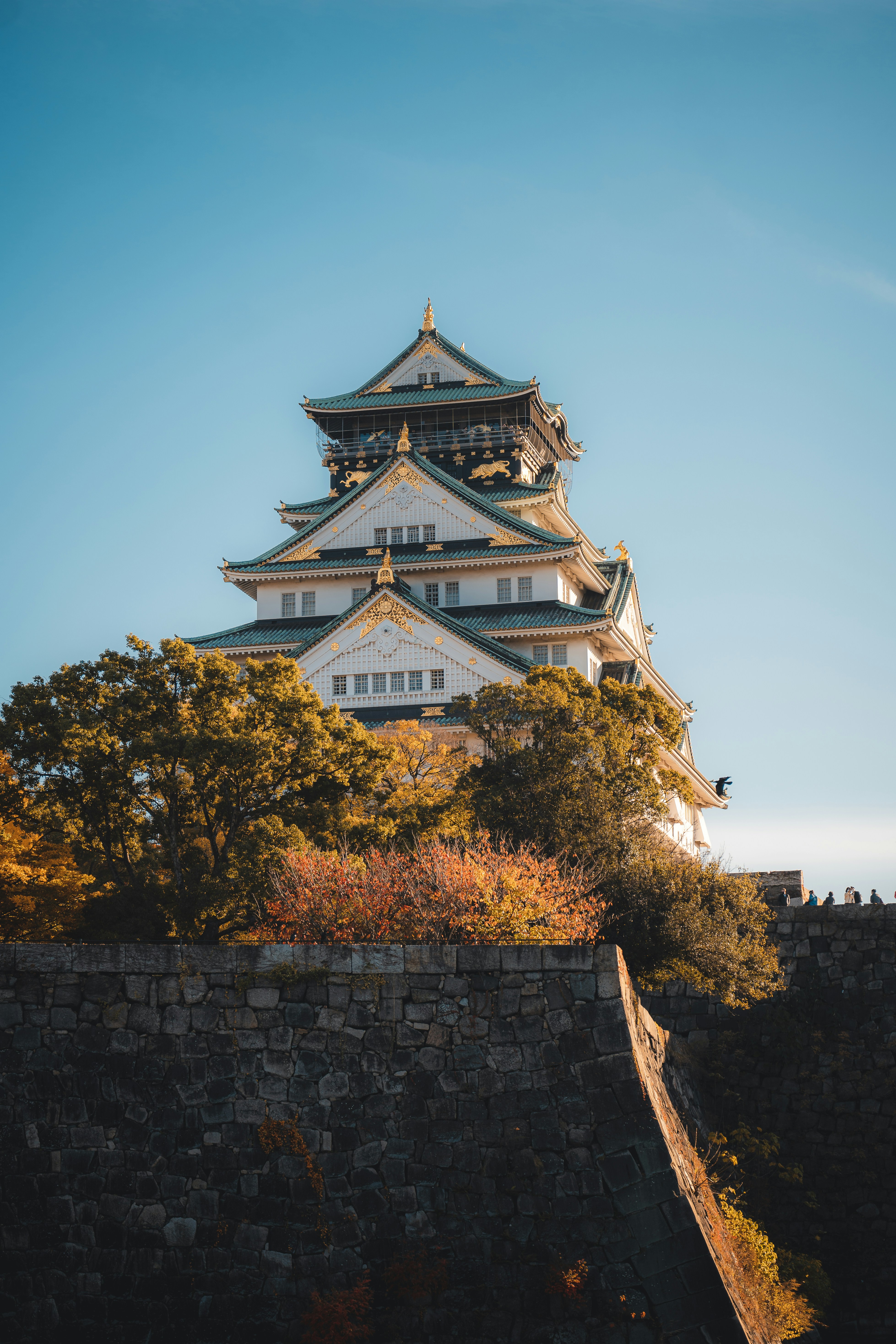 Osaka Castle rises majestically against a clear blue sky, surrounded by vibrant autumn foliage and ancient stone walls.