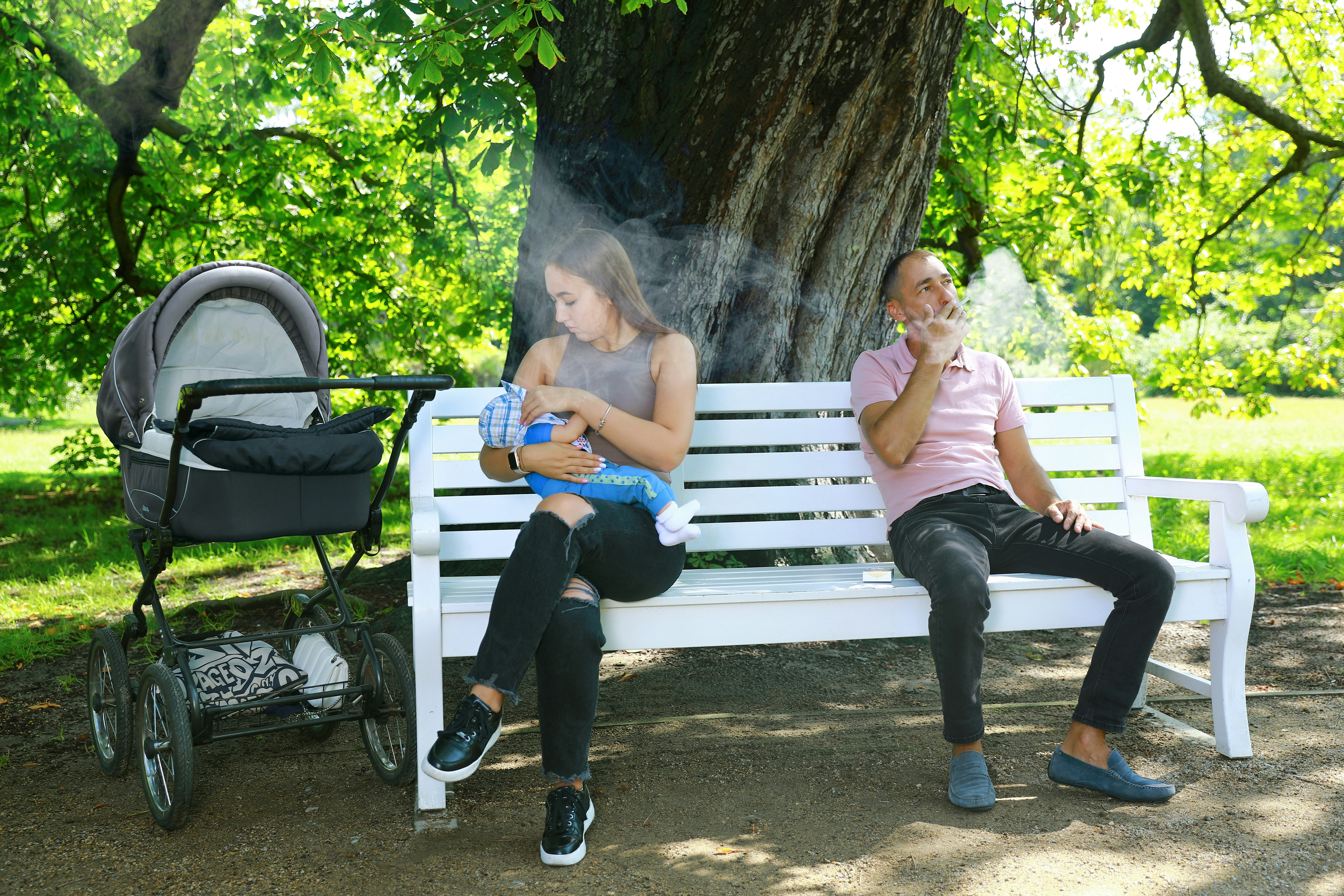 A man smokes a cigarette on a park bench and disturbs a young mother and her baby with his smoke – an appeal for a little more attention and awareness when smoking outdoors. Ein Mann raucht auf einer Parkbank und belästigt mit seinem Rauch eine junge Mutter mit ihrem Baby – ein Aufruf zu etwas mehr Rücksicht und Achtsamkeit beim Rauchen im Freien.