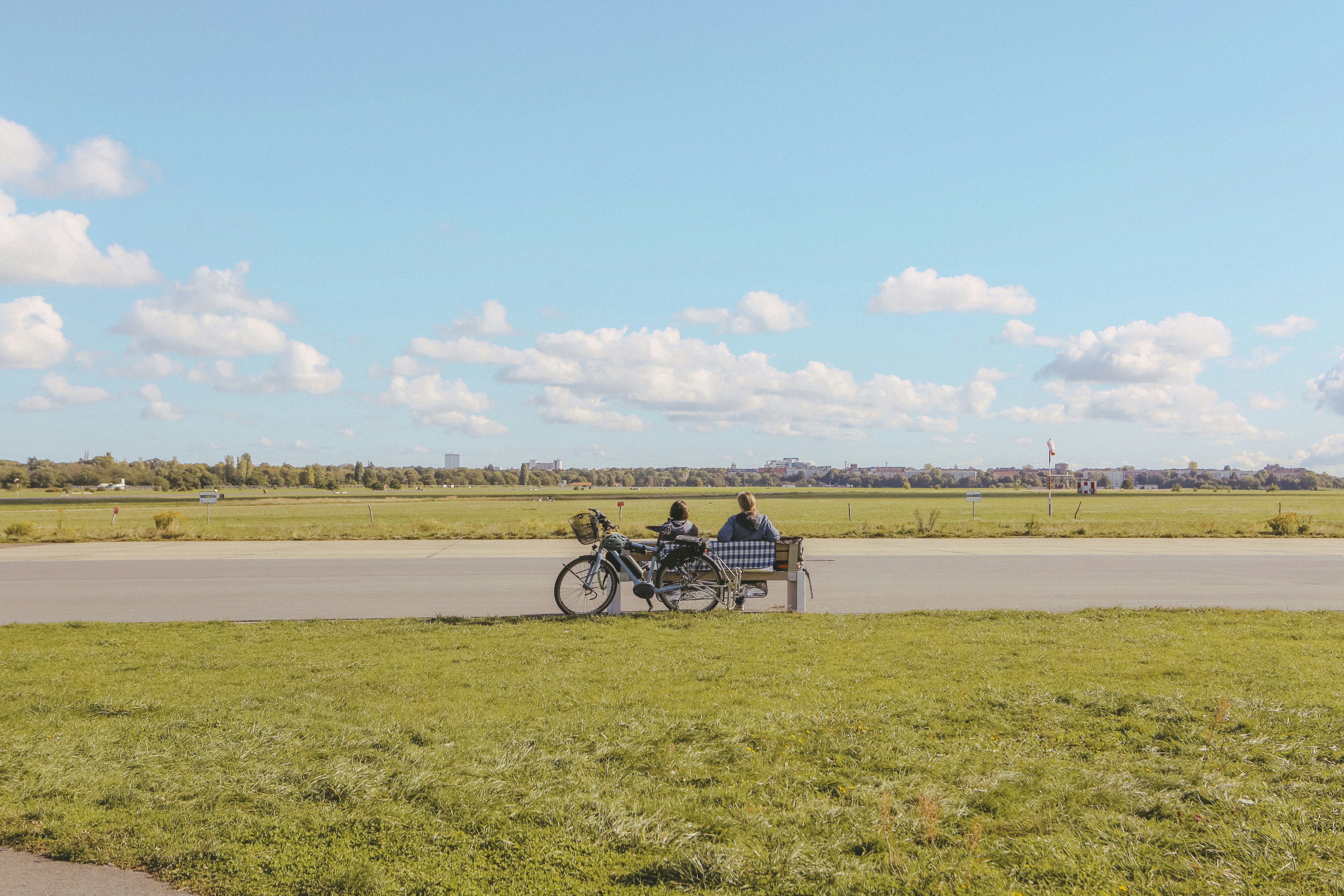 Benchside Pause: Two cyclists rest with their bikes under a sunlit, expansive park sky