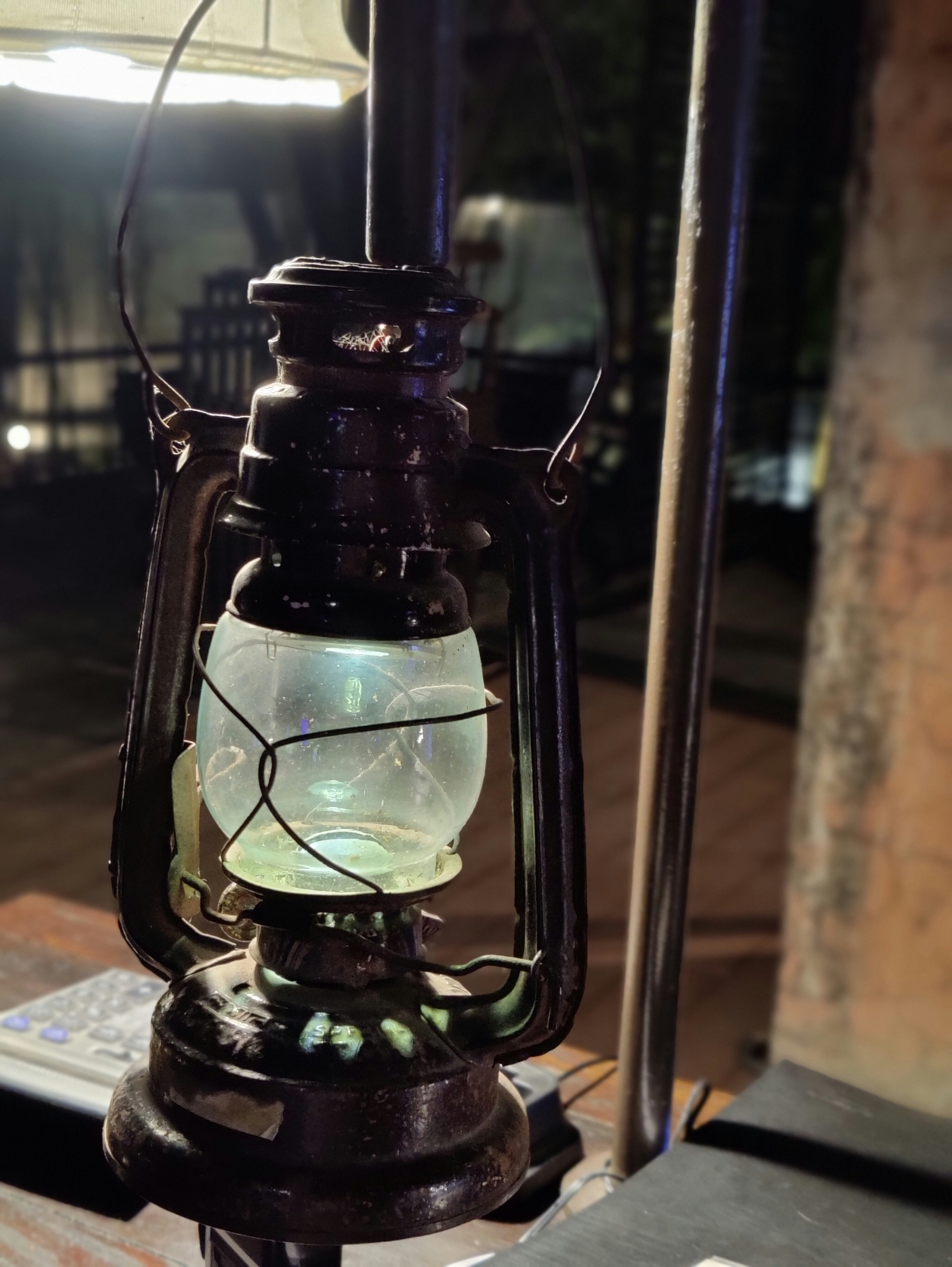 A weathered lantern sits in a dim outdoor setting, its glass globe catching a faint glow. Wires and metal framing create a rustic, industrial feel.