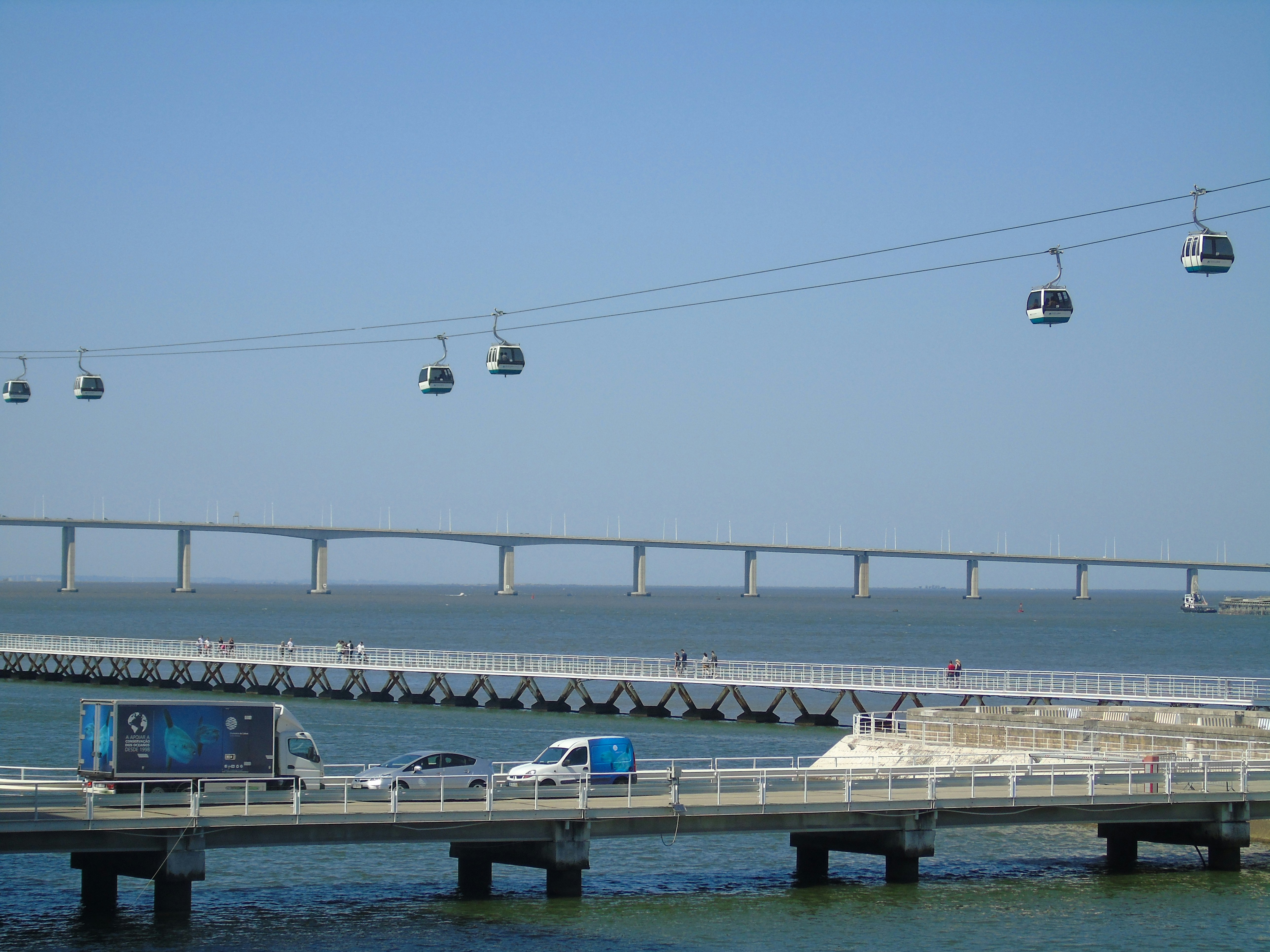 Cable cars glide over a bridge spanning a calm body of water under a clear blue sky.