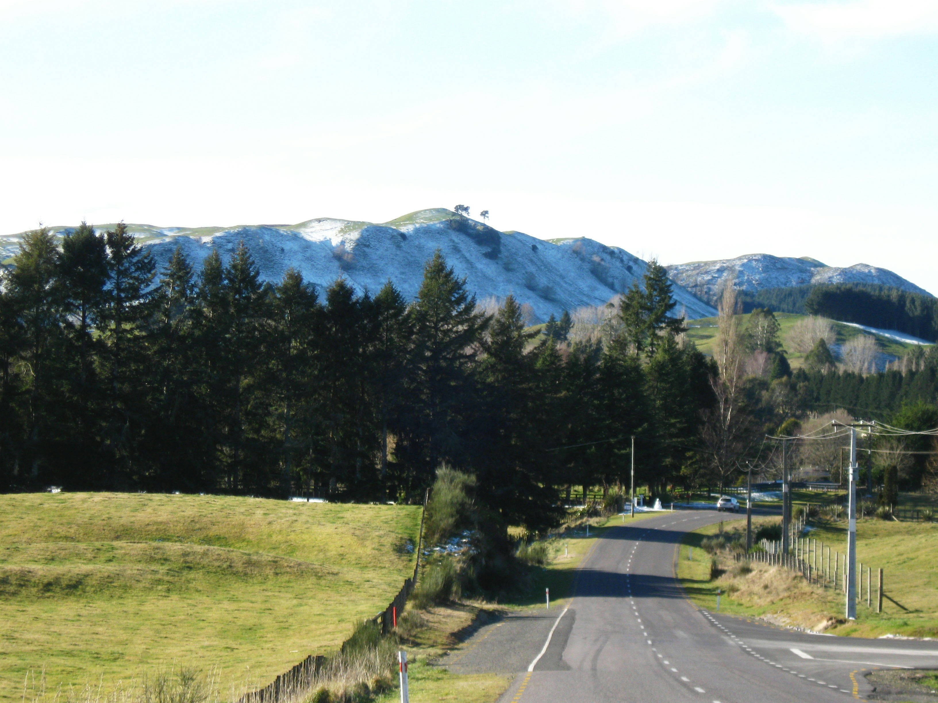 Snow-dusted hills rise behind a winding road, framed by lush green fields and dense forests. The scene captures the tranquil beauty of a winter day.