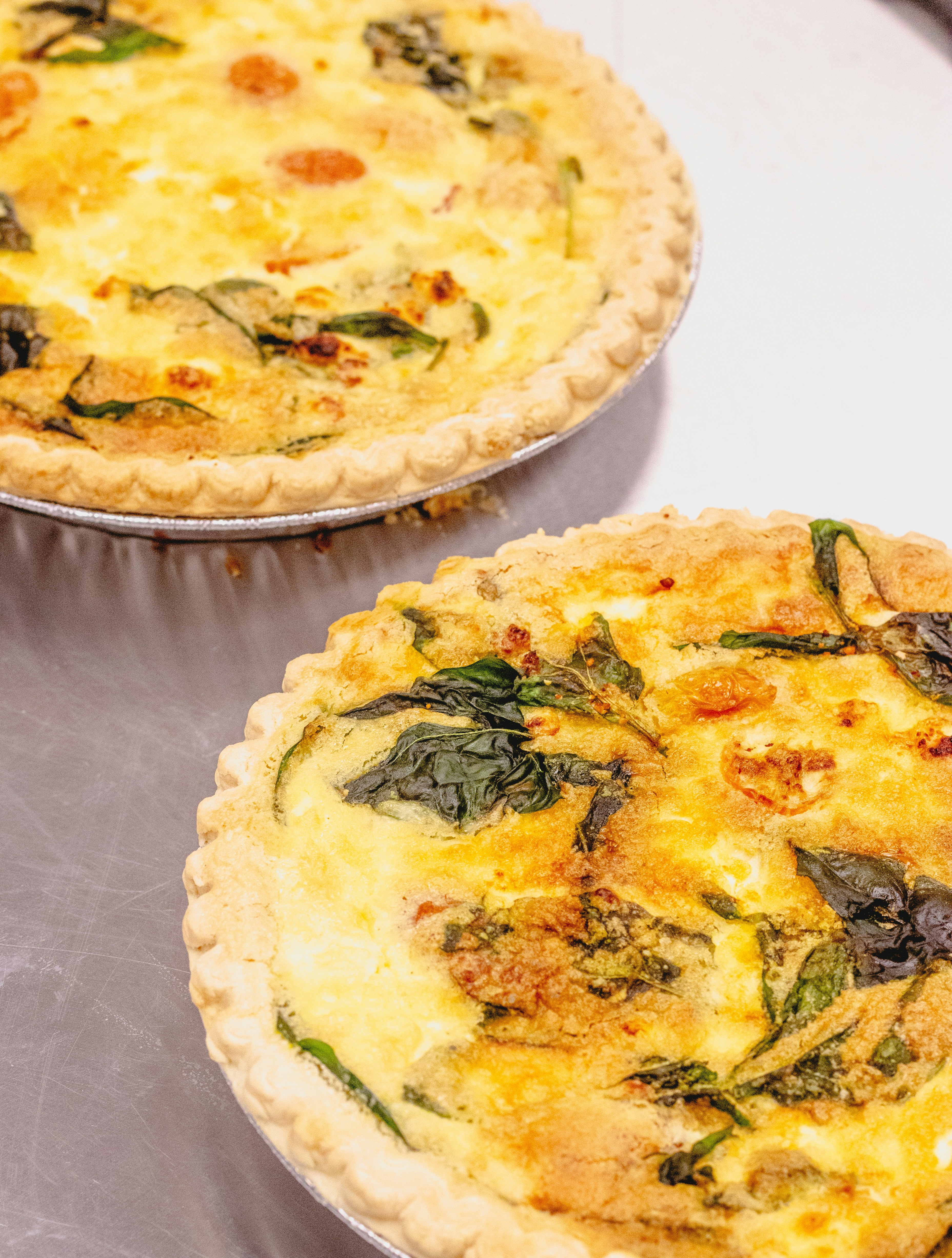 Two freshly baked quiches featuring vibrant spinach and sun-dried tomatoes, resting on a stainless steel surface.
