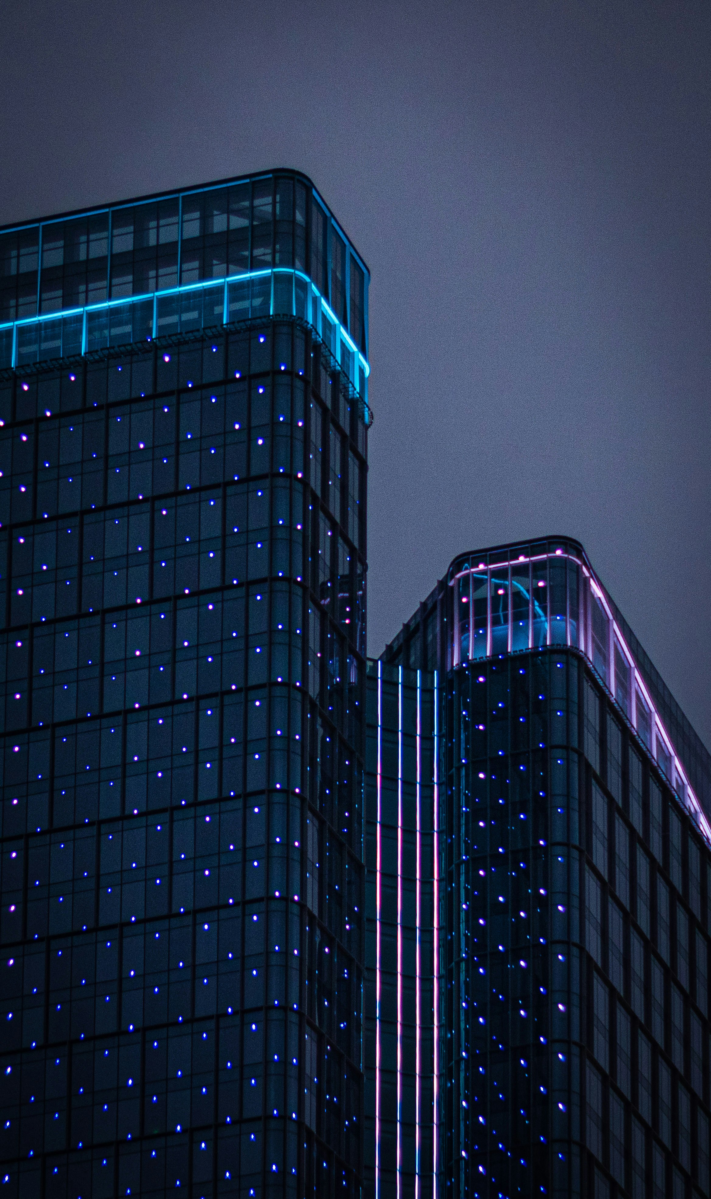 Illuminated glass towers with vibrant blue and pink lighting against a twilight sky. The interplay of colors and reflections highlights the building's sleek design.