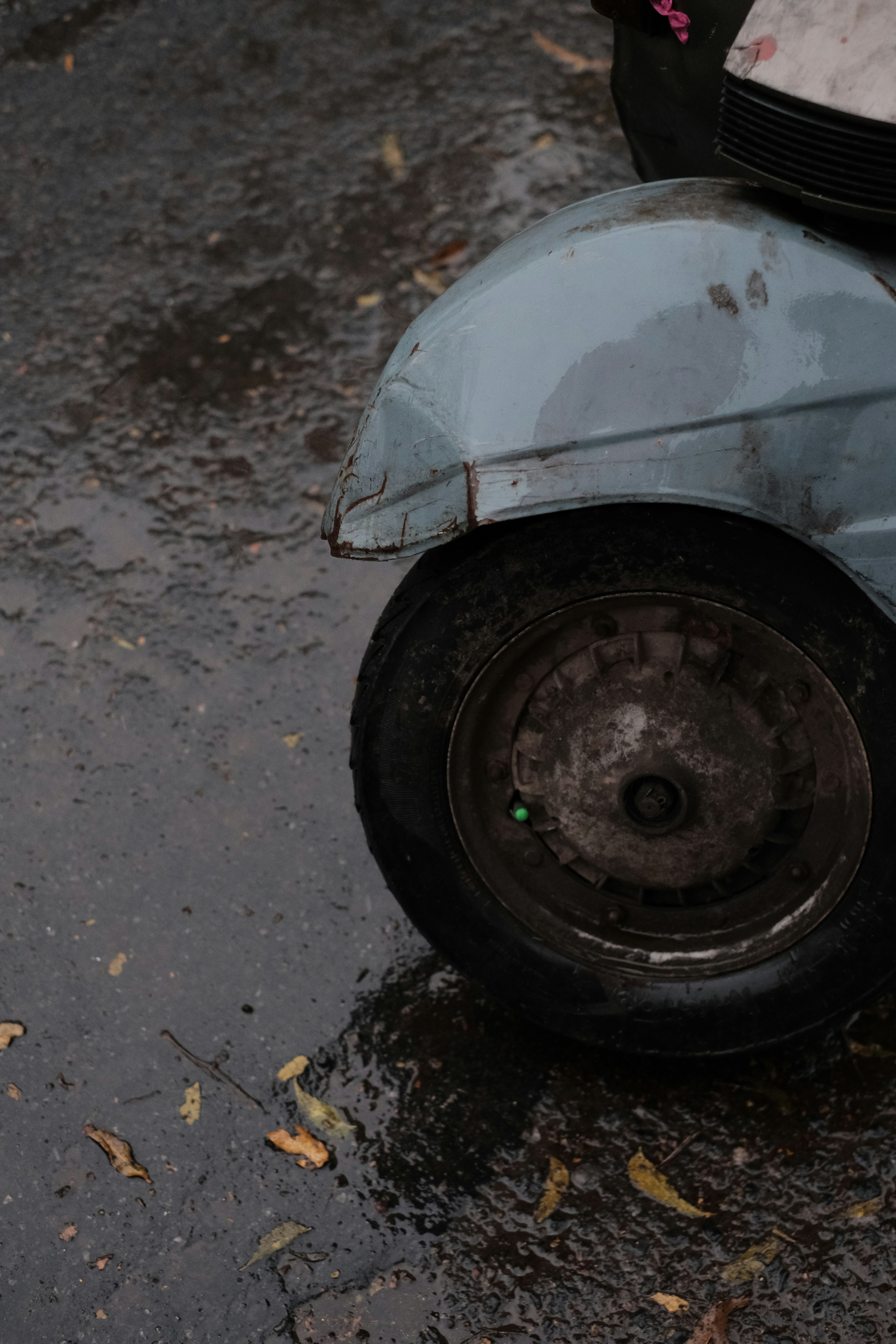 Weathered blue scooter fender frames a rusted wheel on rain-soaked pavement