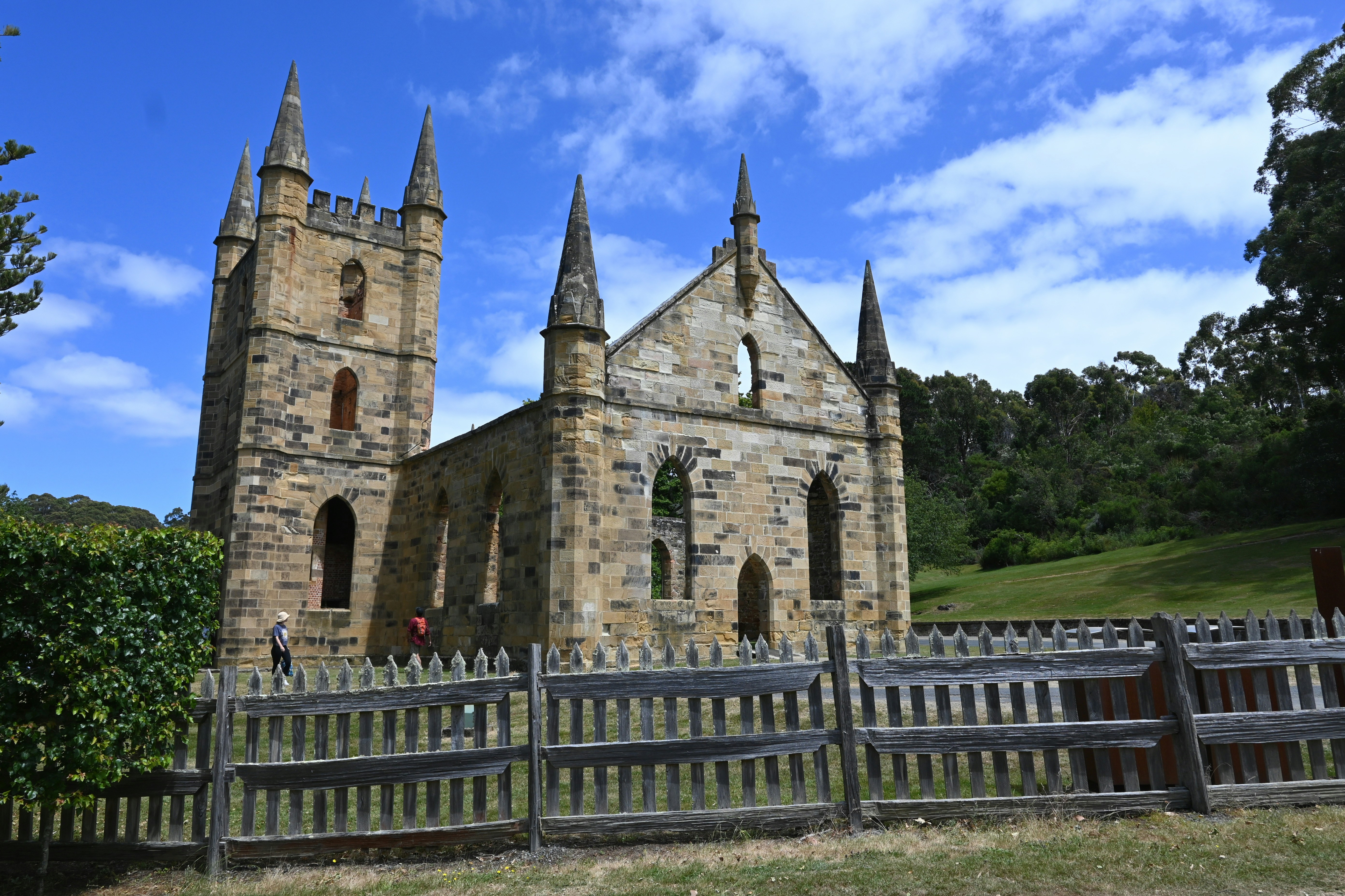 Stone cathedral ruins with towering spires stand behind a wooden fence under a bright blue sky.