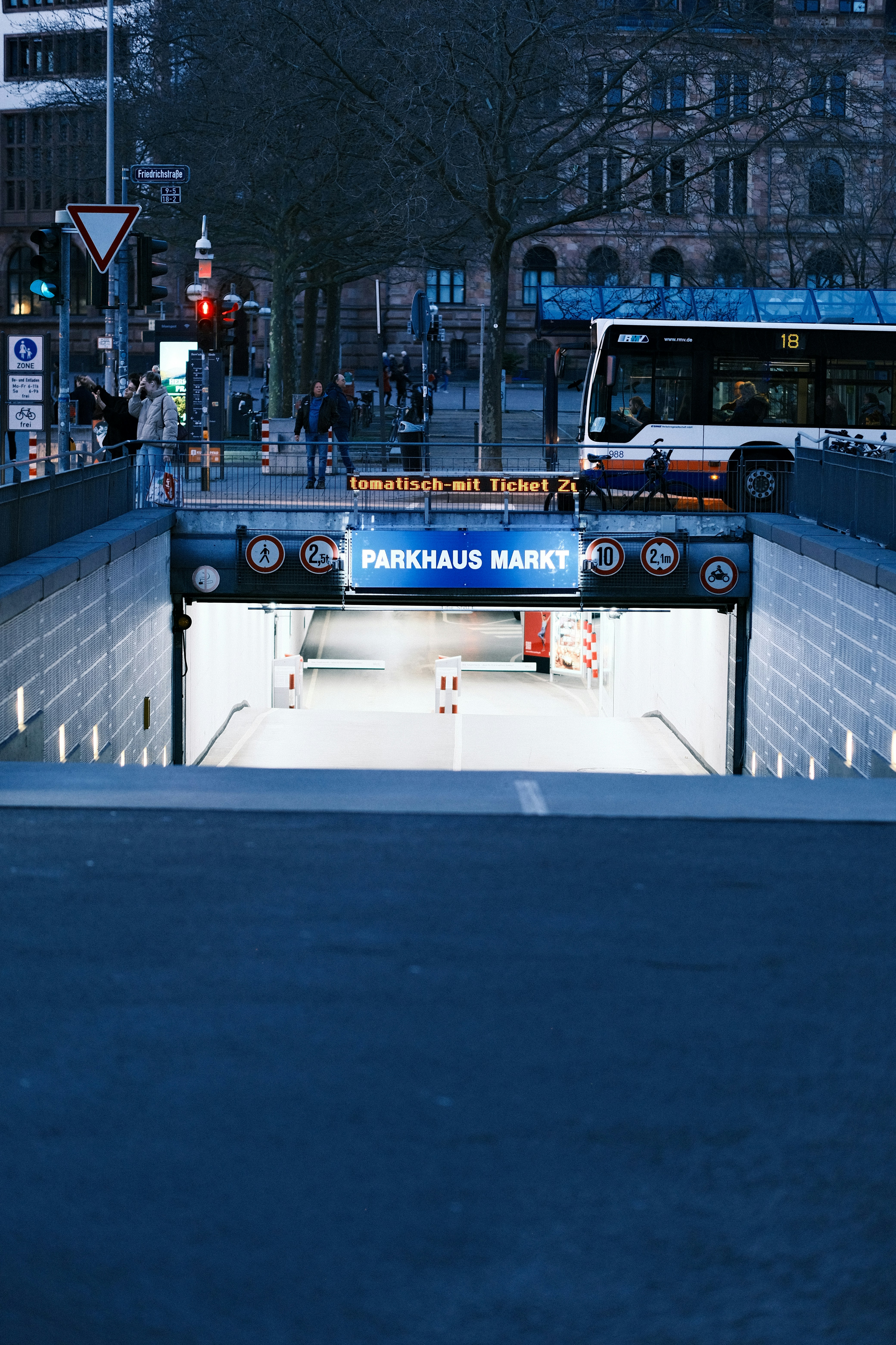 Entrance to an underground parking garage marked by a glowing sign, surrounded by pedestrians and urban architecture.