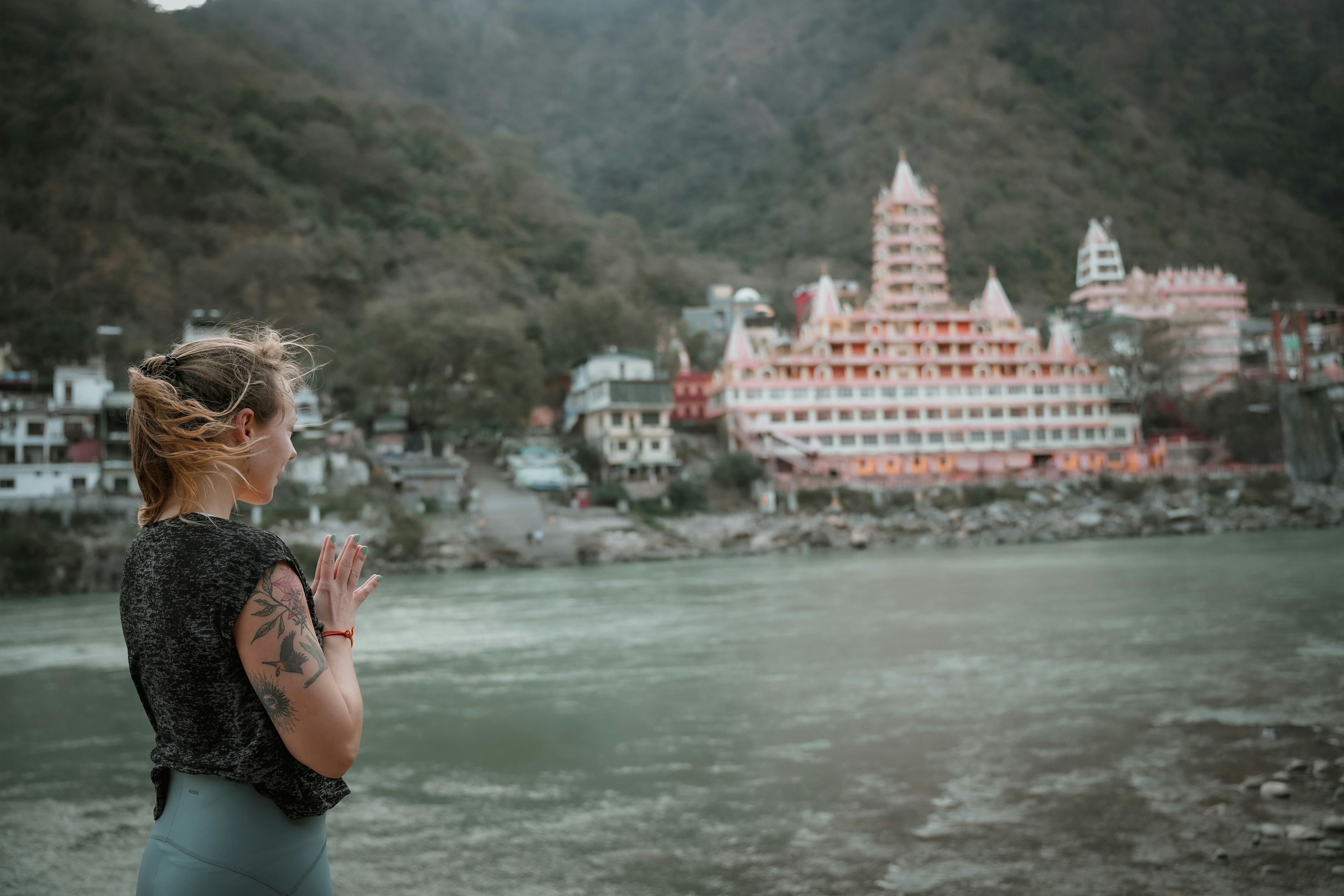 A woman with a tattooed arm stands in prayer by the river, gazing at a beautifully lit temple across the water. The serene landscape captures the essence of spirituality and tranquility.