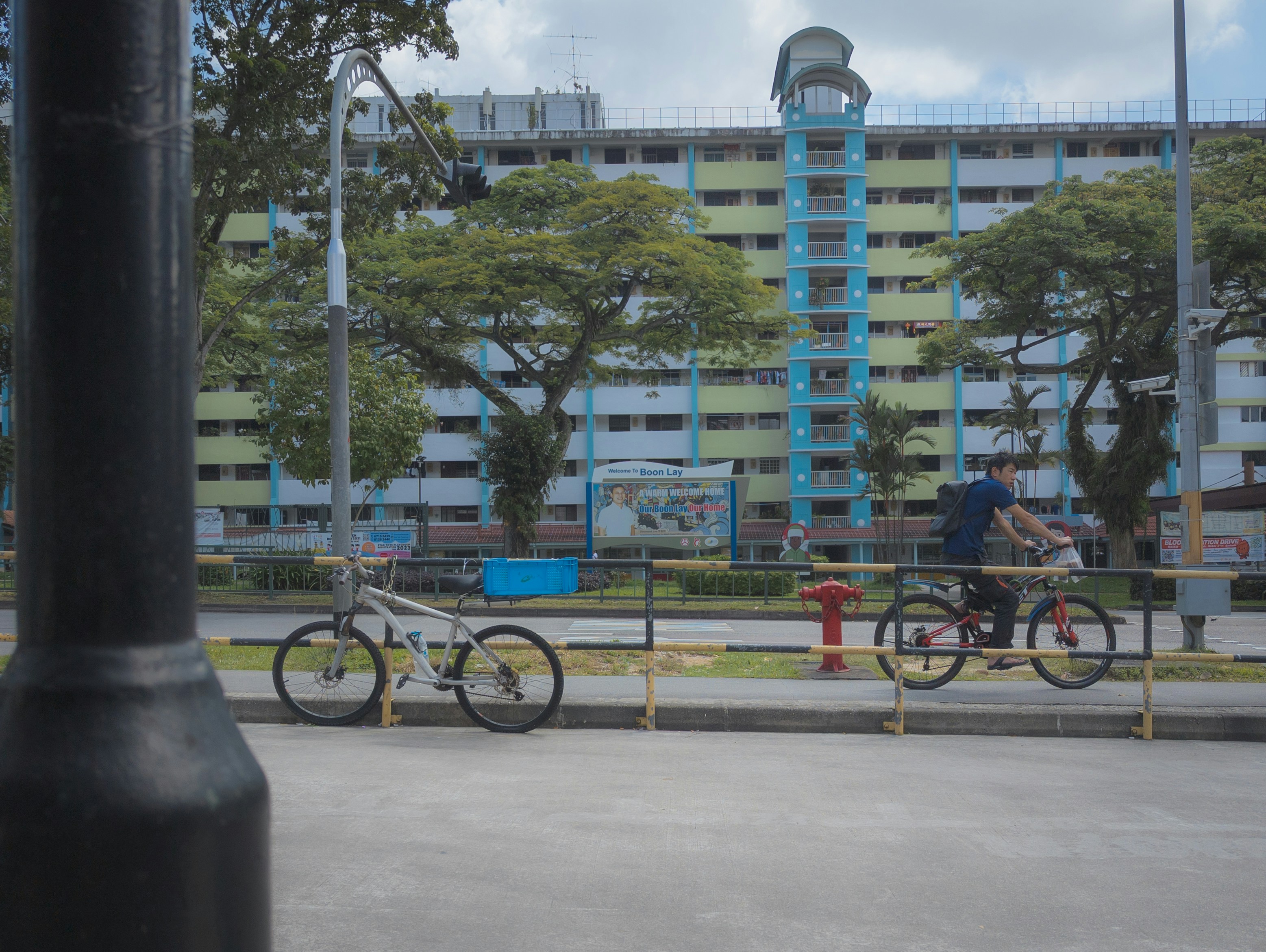 Two cyclists navigating a vibrant urban landscape, flanked by a colorful apartment building and lush trees. The scene captures the essence of everyday life in the city.