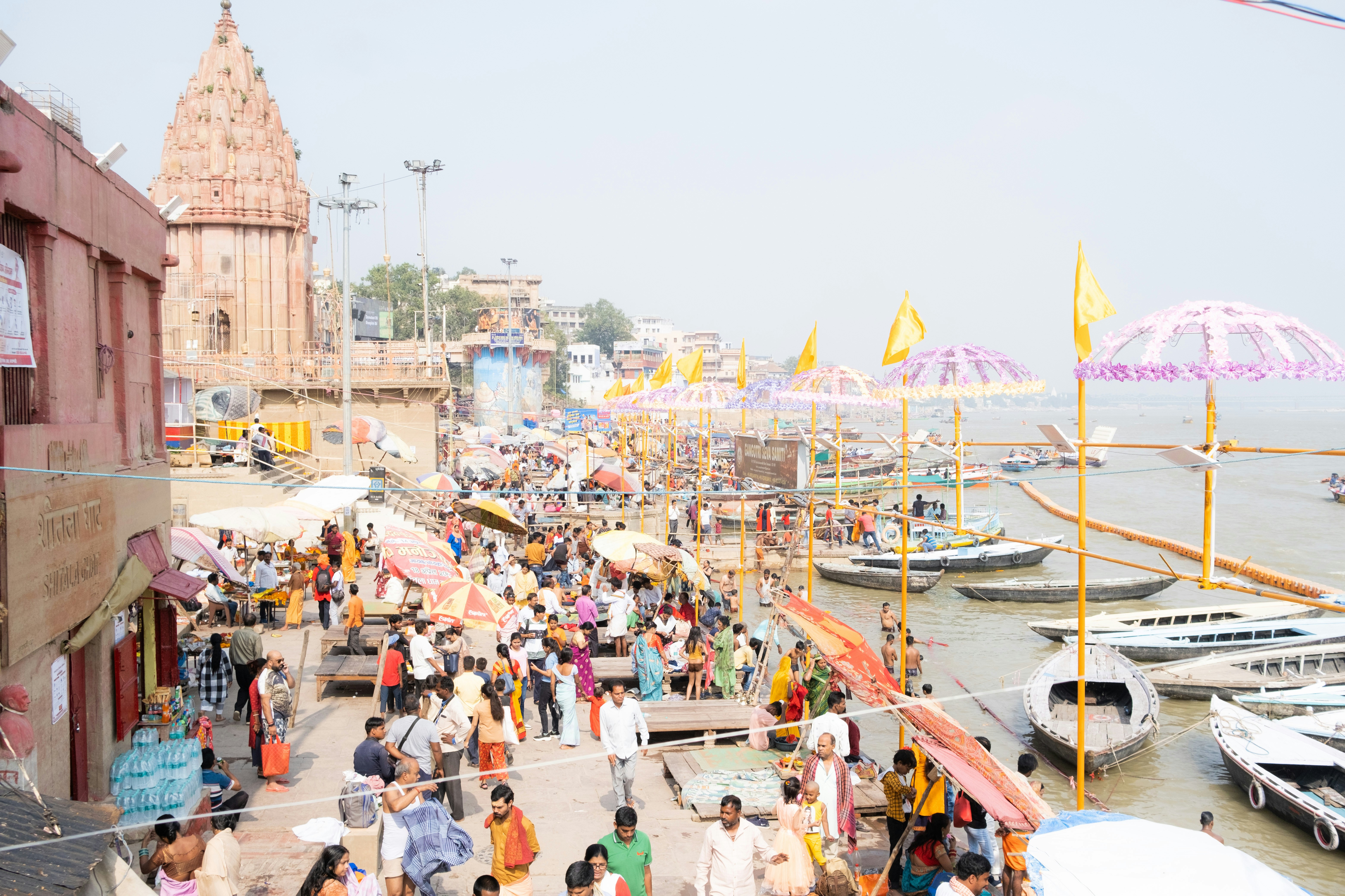Crowded riverside ghats with people, boats, and colorful umbrellas under a hazy sky.
