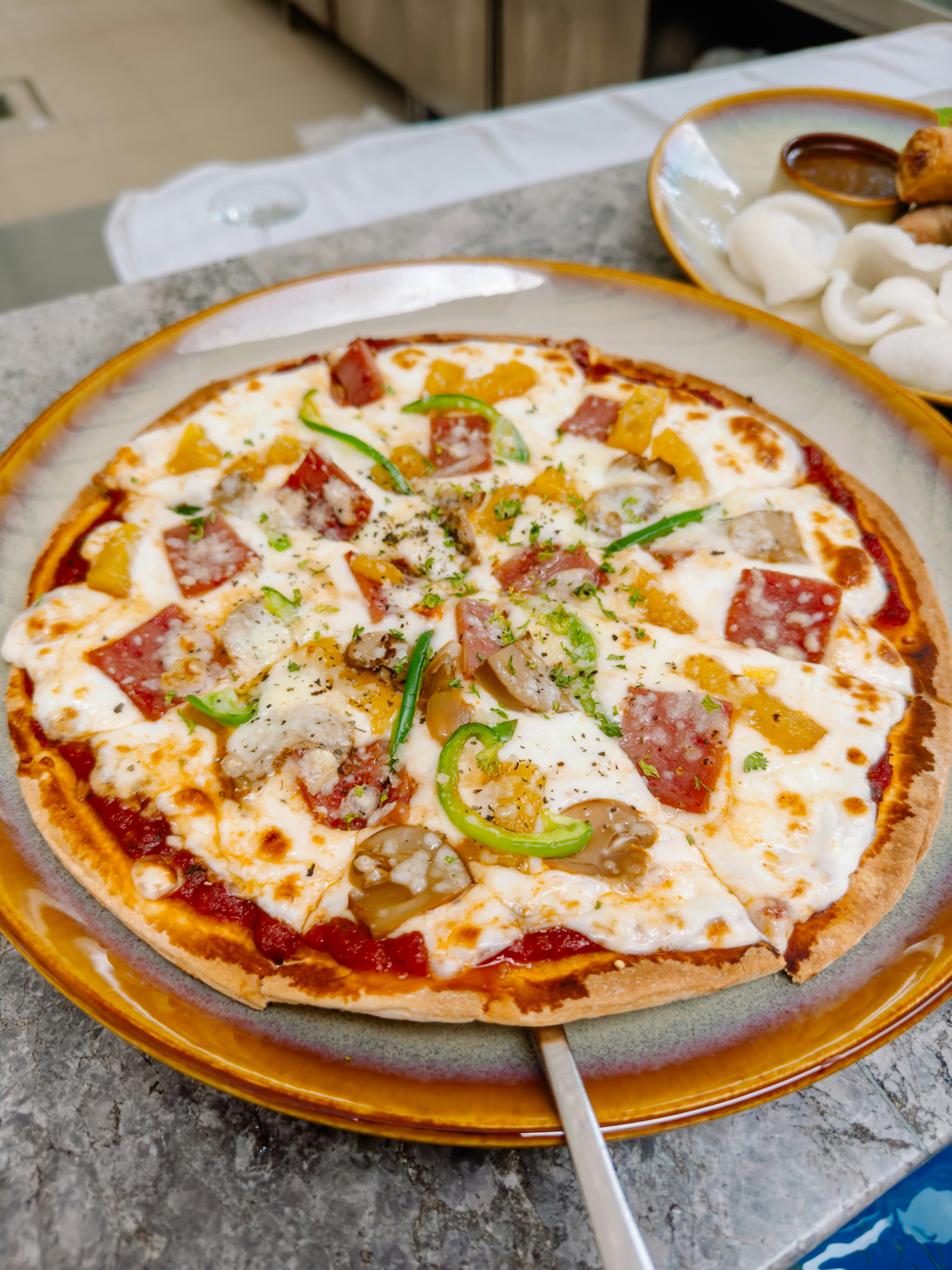 Cheesy pizza topped with pepperoni, bell peppers, and mushrooms sits on a warm plate with a side of bread; a casual dining scene at a marble counter.