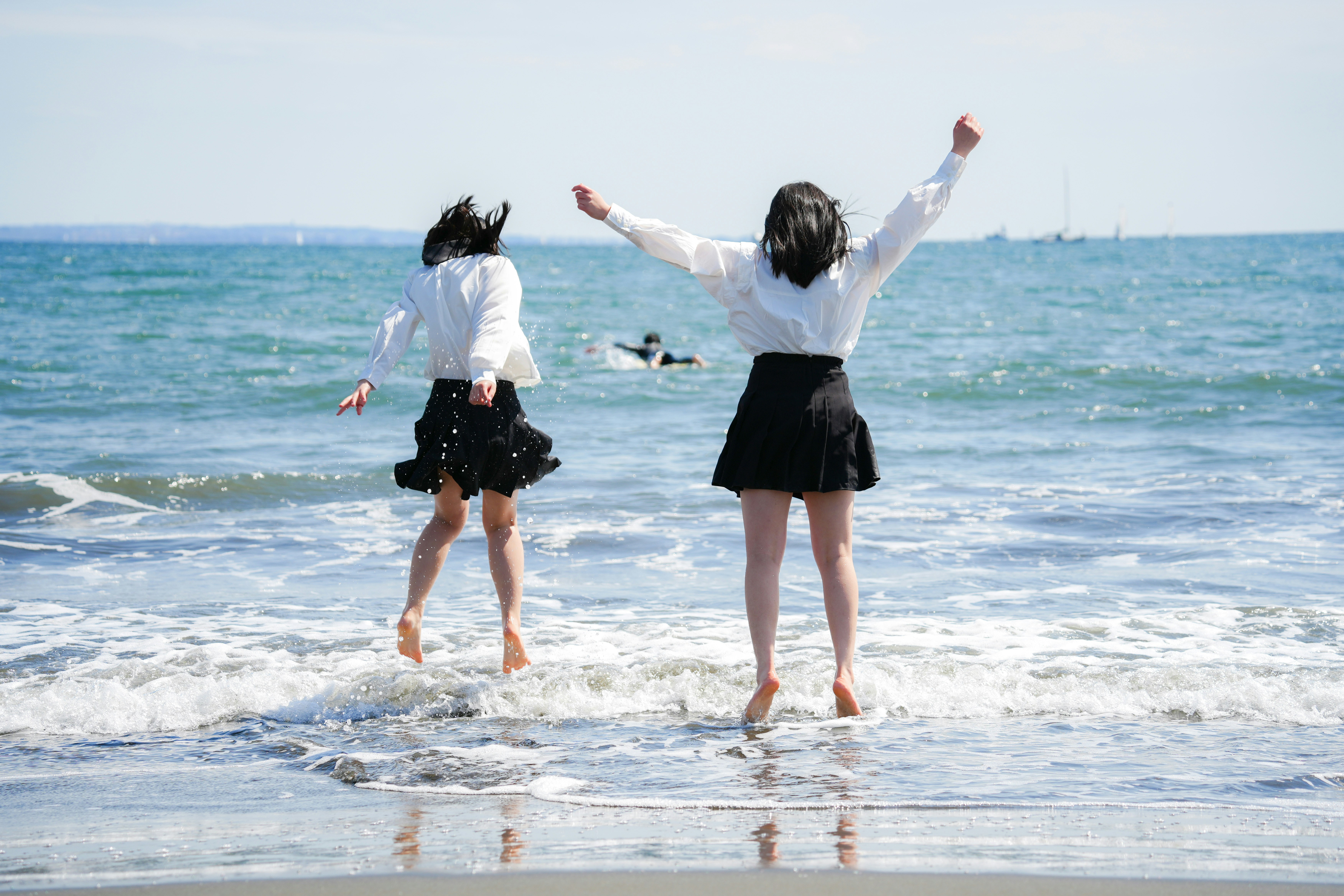Two women in black skirts and white tops jump playfully along the shoreline with waves crashing around them.