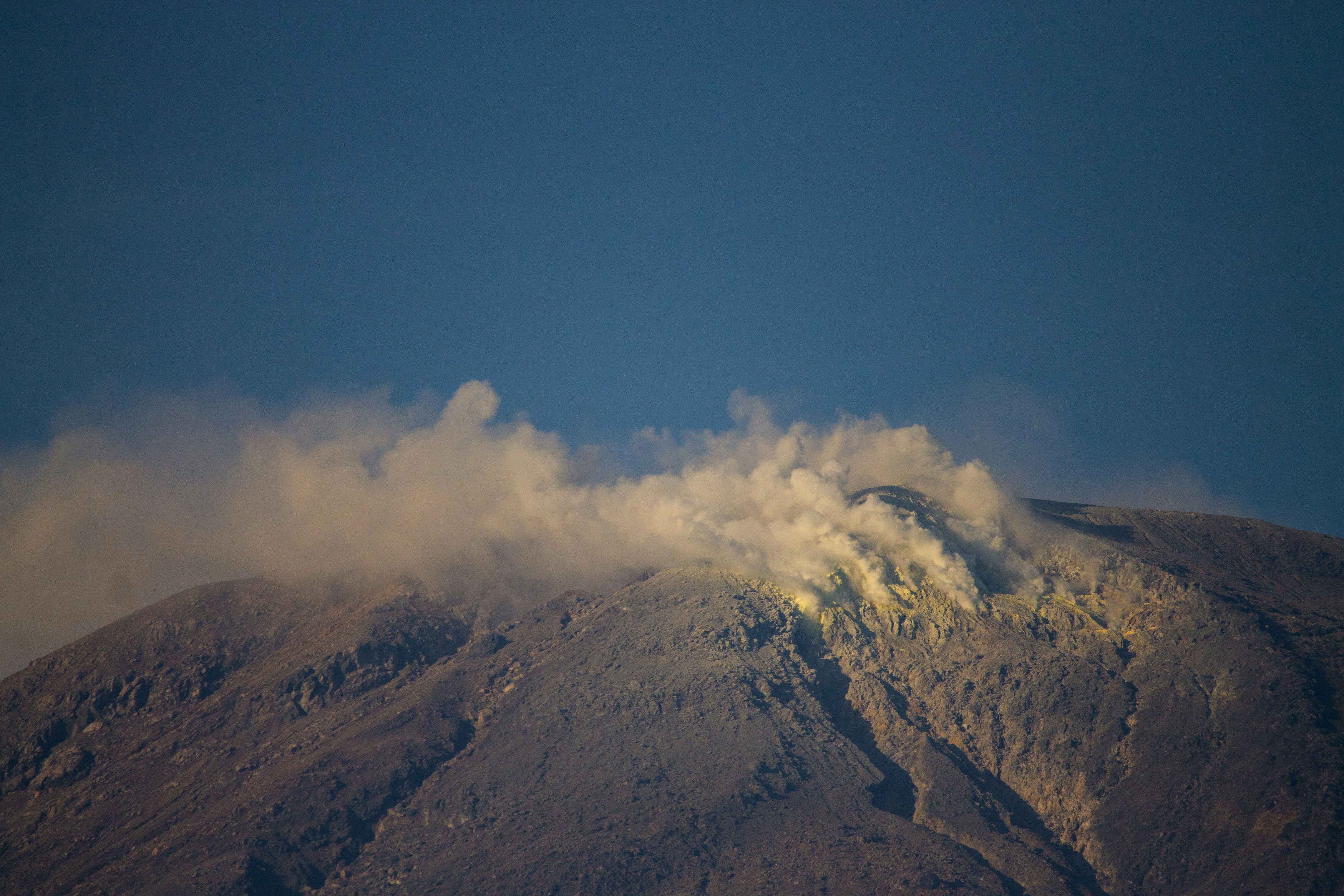 Volcanic smoke drifts from a rugged mountain peak against a clear blue sky.