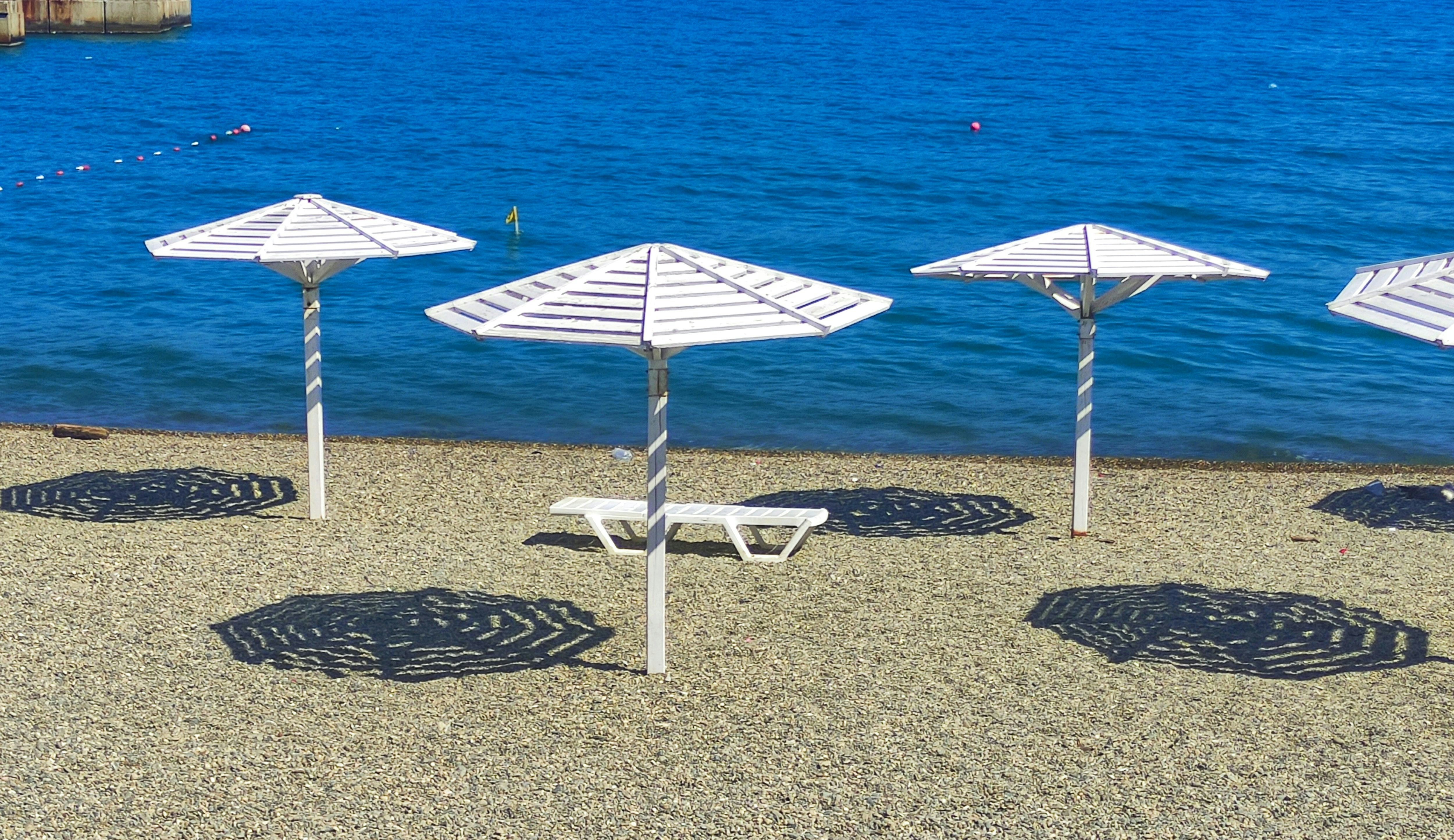 White umbrellas casting intricate shadows on a sandy beach beside a calm blue sea.