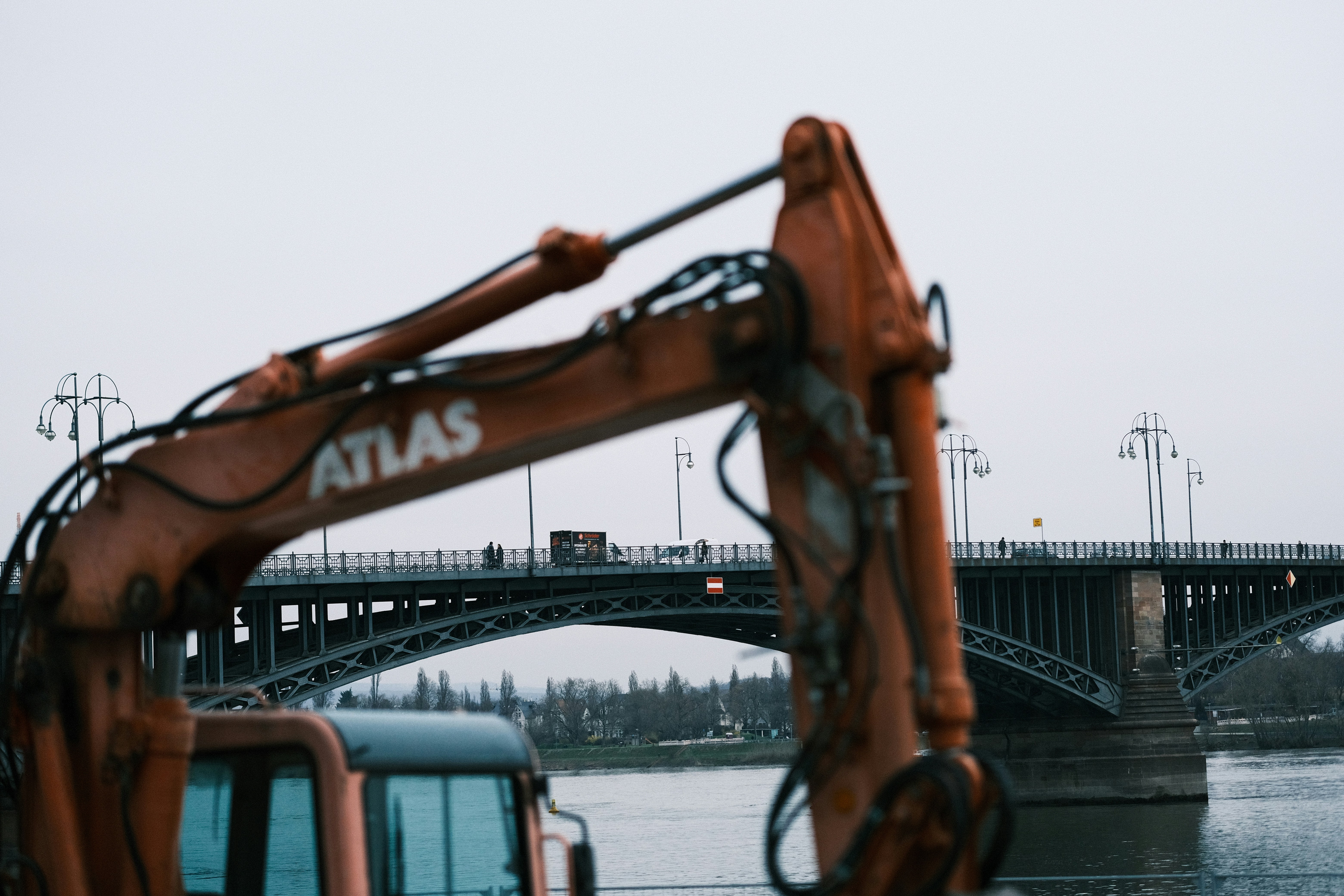 Excavator arm frames a distant bridge over a calm river under an overcast sky.